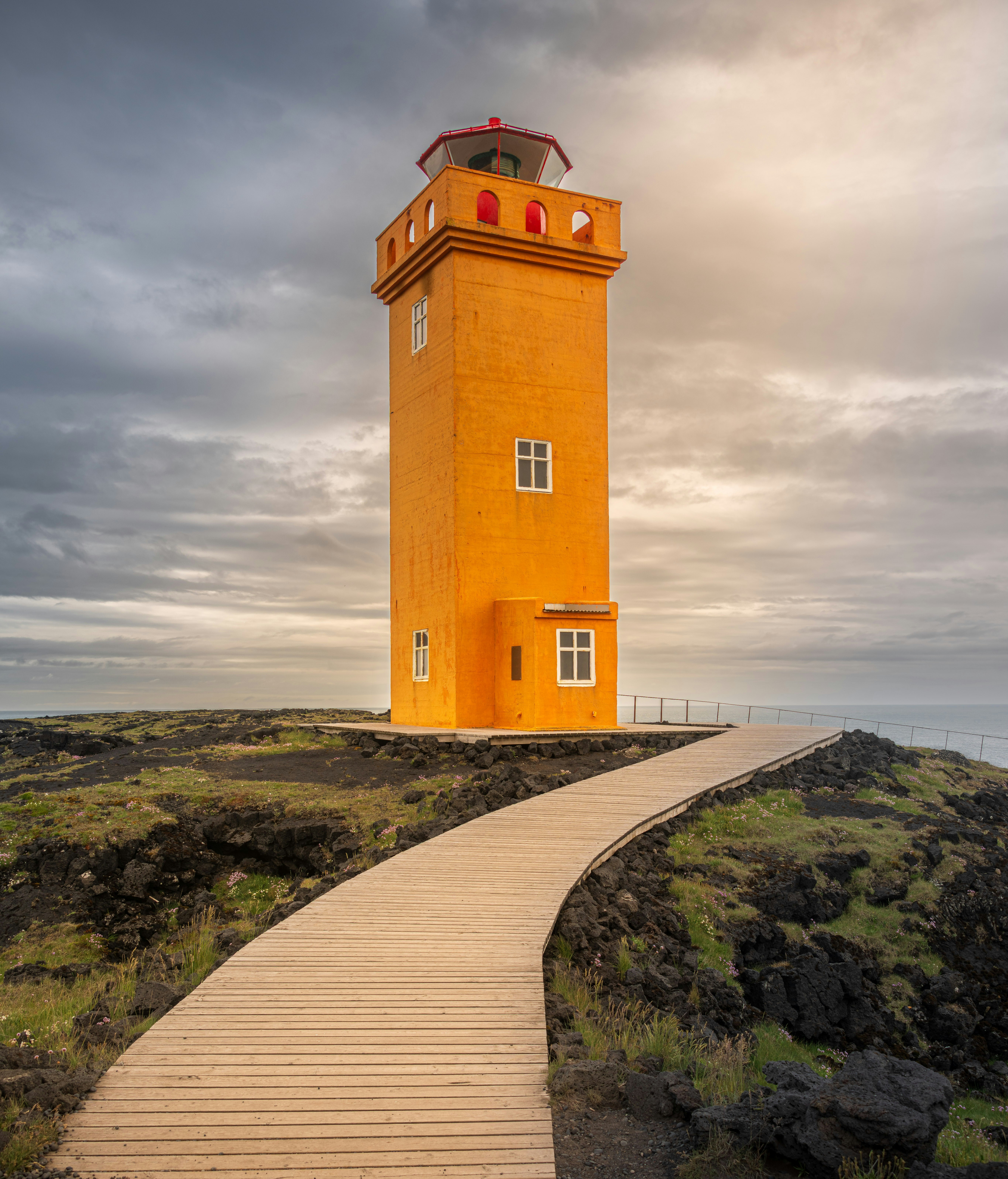 The vivid orange Svortuloft lighthouse rises from Iceland's black lava fields on the Snæfellsnes coast, reached by a winding boardwalk beneath brooding Icelandic skies. | Orange lighthouse on a rocky coast with wooden path