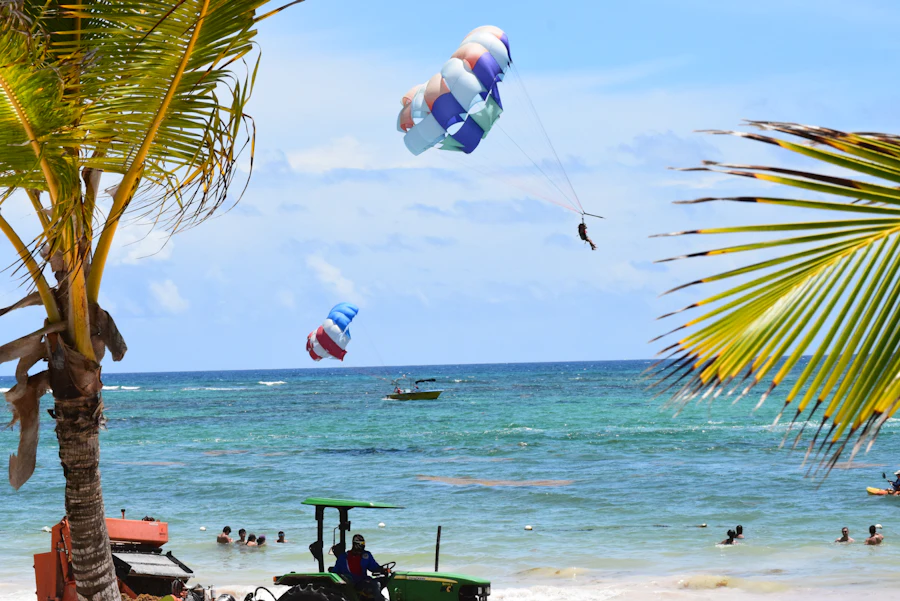 Two parasailers drift above a bright turquoise sea, framed by palm fronds on both sides. Swimmers, a small boat, and a beach vehicle add activity along the shoreline.