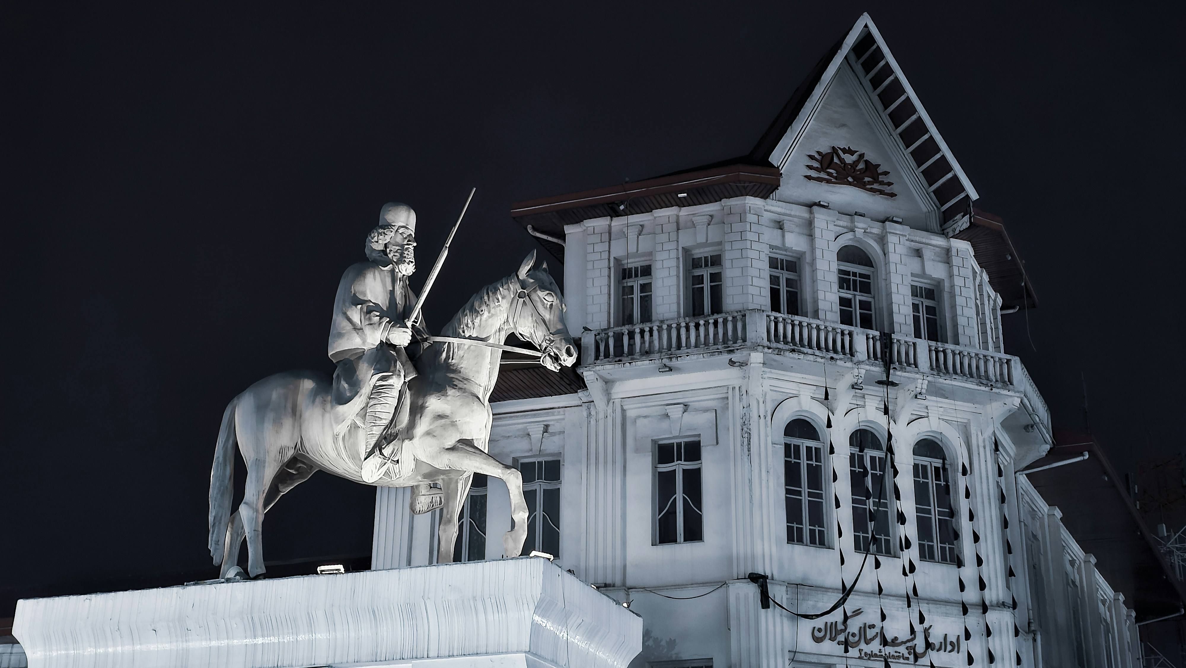 In the heart of Rasht. Rasht’s Shahrdari Square at night – where history and identity meet. The statue of Mirza Kuchak Khan Jangali, a legendary freedom fighter who led the Jangal movement in early 20th-century Iran, stands proudly in front of the historic Post Office building. Together, they tell a story of resistance, heritage, and the cultural richness of Gilan. This night scene reveals more than architecture; it reflects the enduring memory of those who stood for justice and the city that keeps their spirit alive. | Equestrian statue in front of ornate building at night