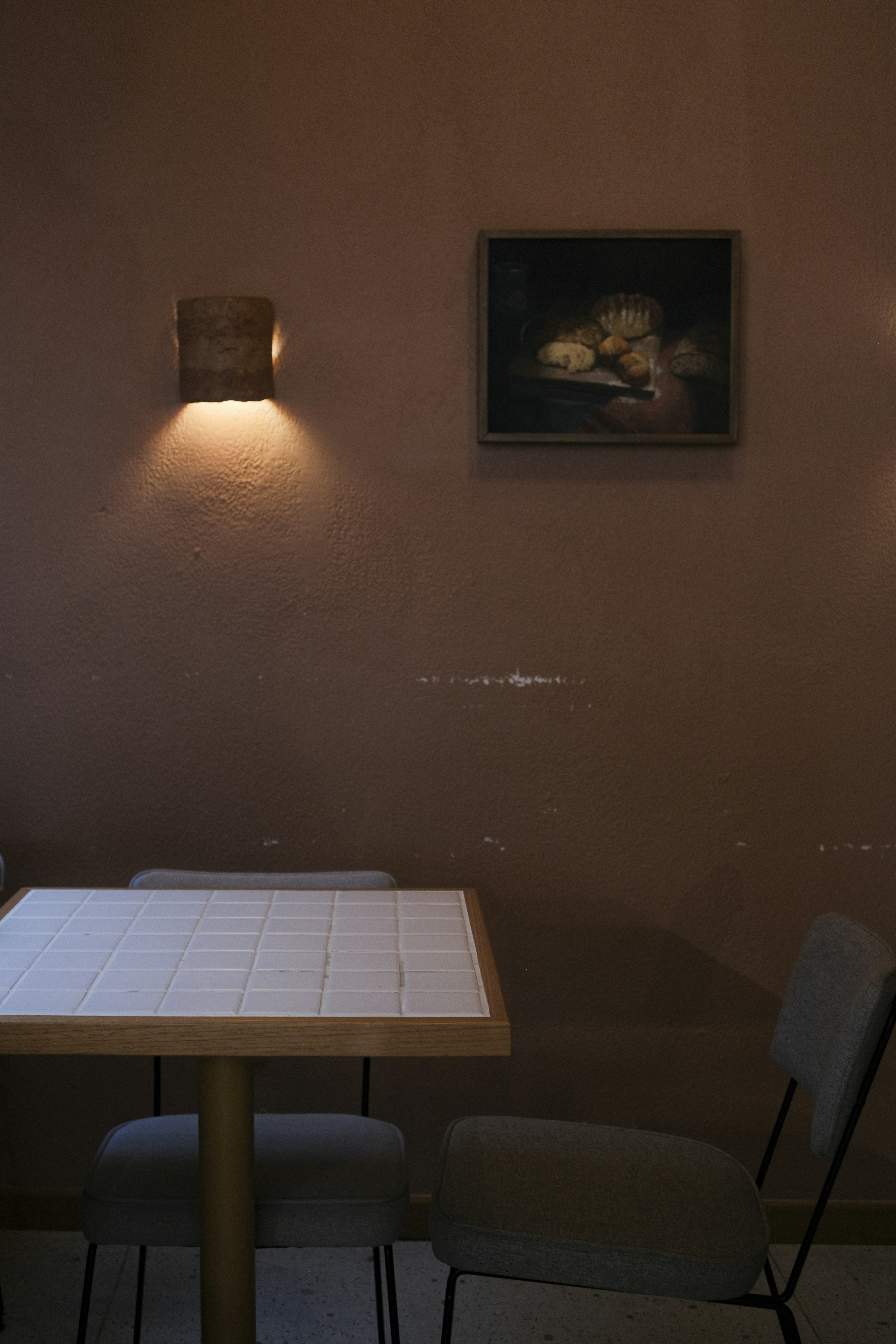 Minimalist dining area featuring a wooden table and gray chairs, illuminated by soft wall sconces, with a still life painting of baked goods on the wall.