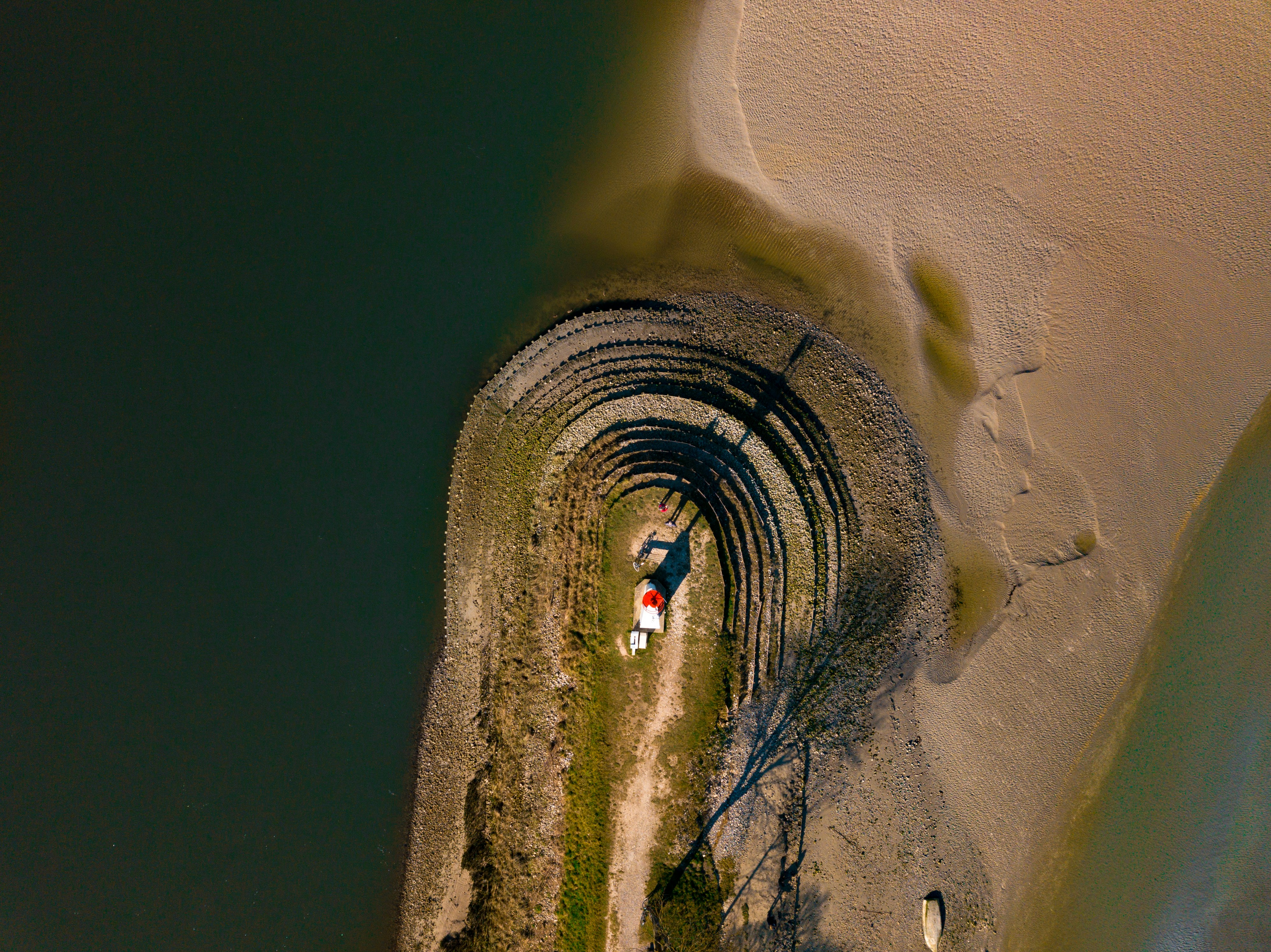 Aerial view of a curved pathway leading to a small structure on a riverbank, surrounded by contrasting textures of land and water.