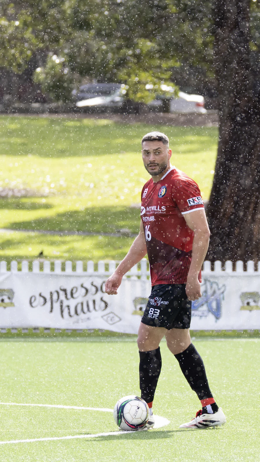 Soccer player in a red jersey standing over the ball on a rain-speckled pitch, gaze directed toward play.