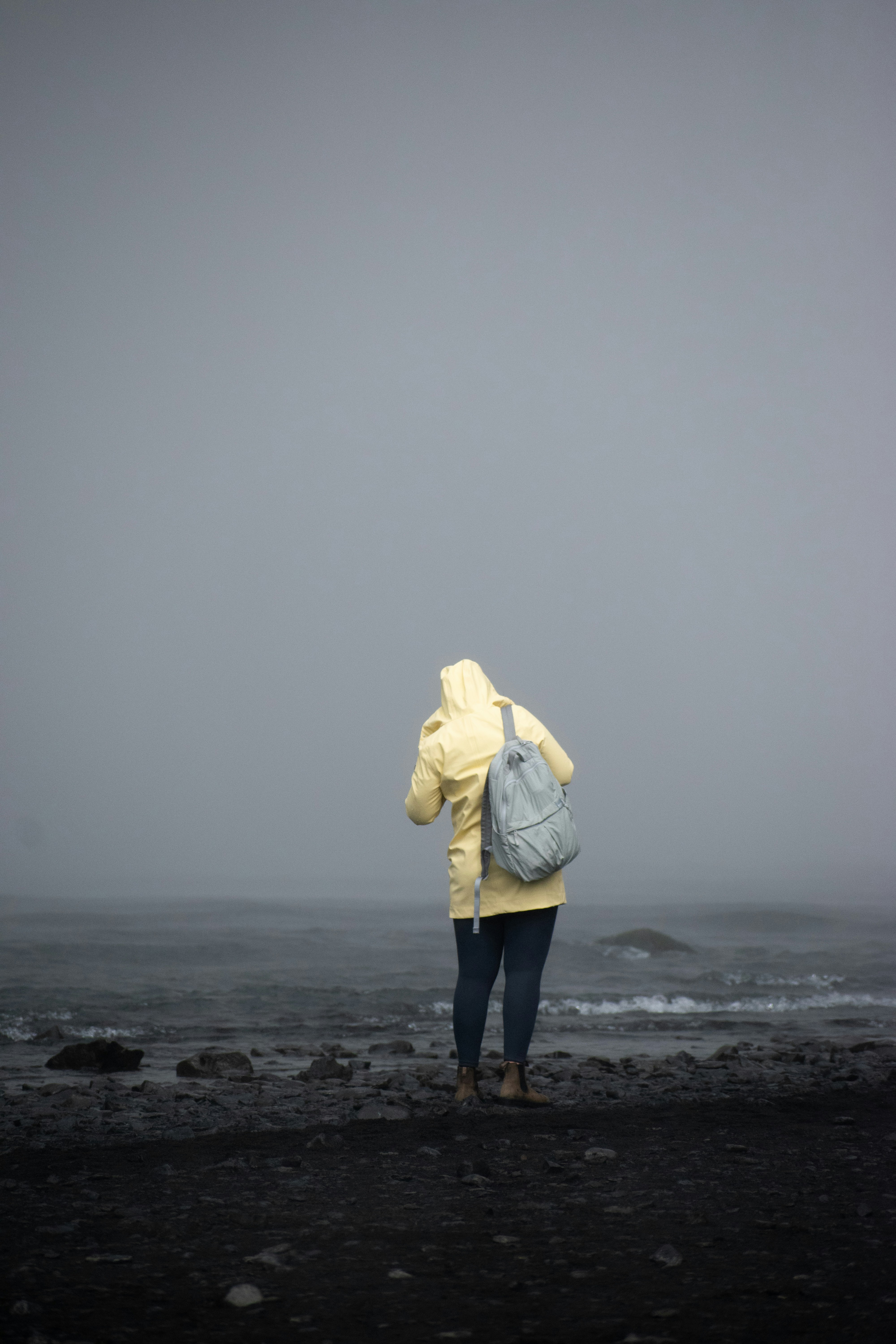 A woman, standing along in the mist of a waterfall | Person in yellow raincoat stands by misty ocean shore.