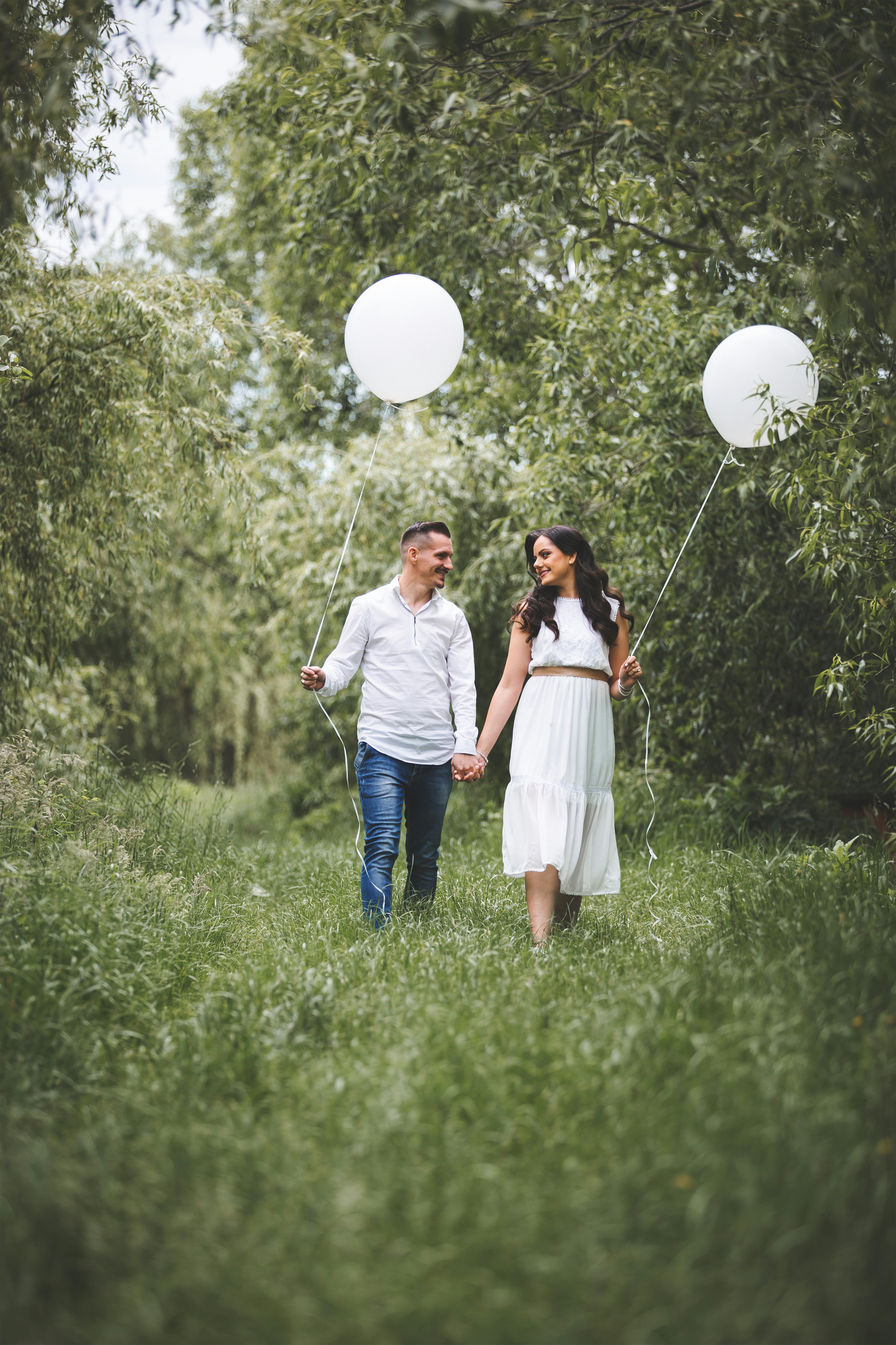 Man and his fiancé walking through grass, holding balloons in their hands looking at each other and smiling. | Couple walking in a grassy park holding balloons