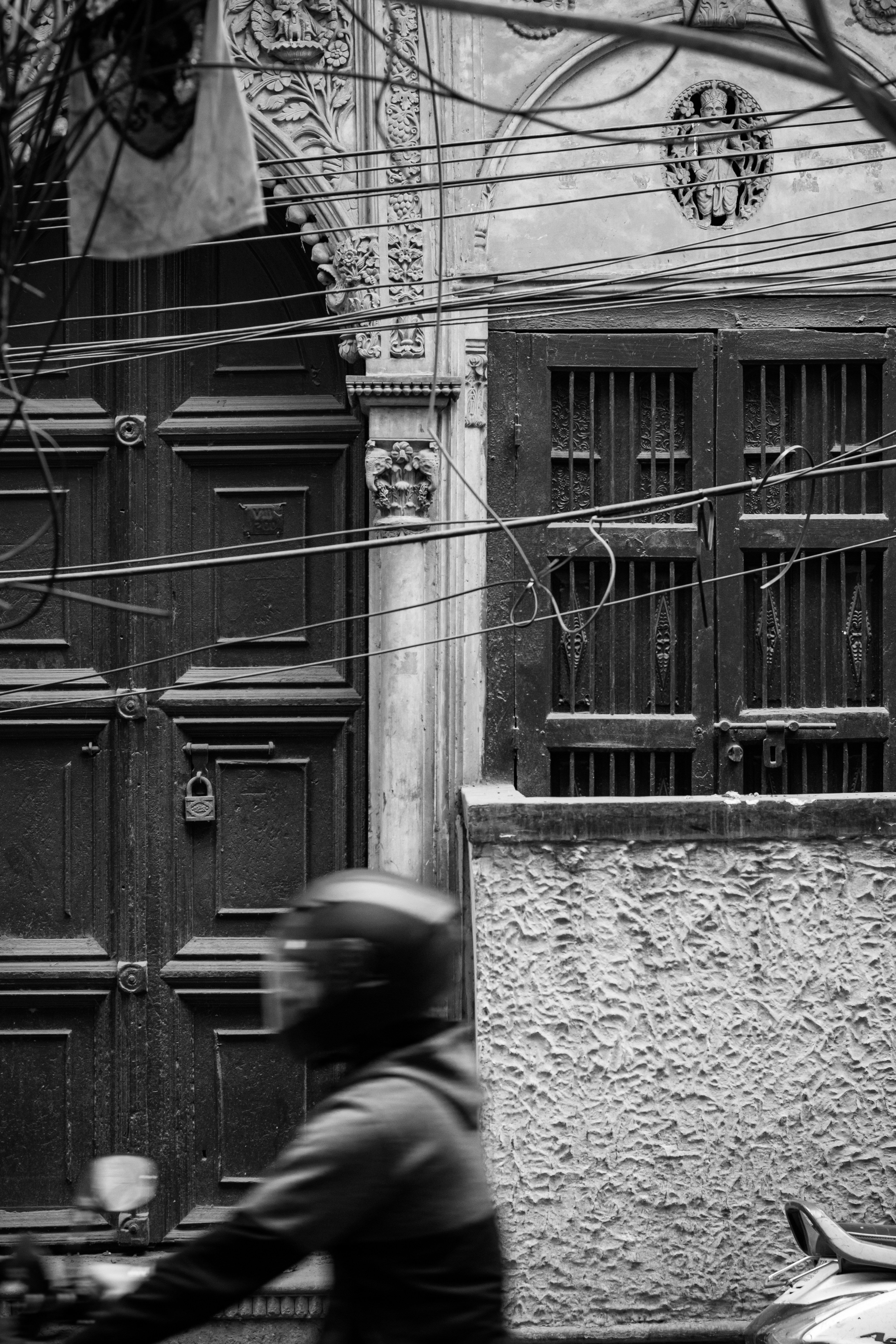 A motorcyclist speeds past intricately detailed doors, framed by a web of overhead wires in a bustling urban setting.