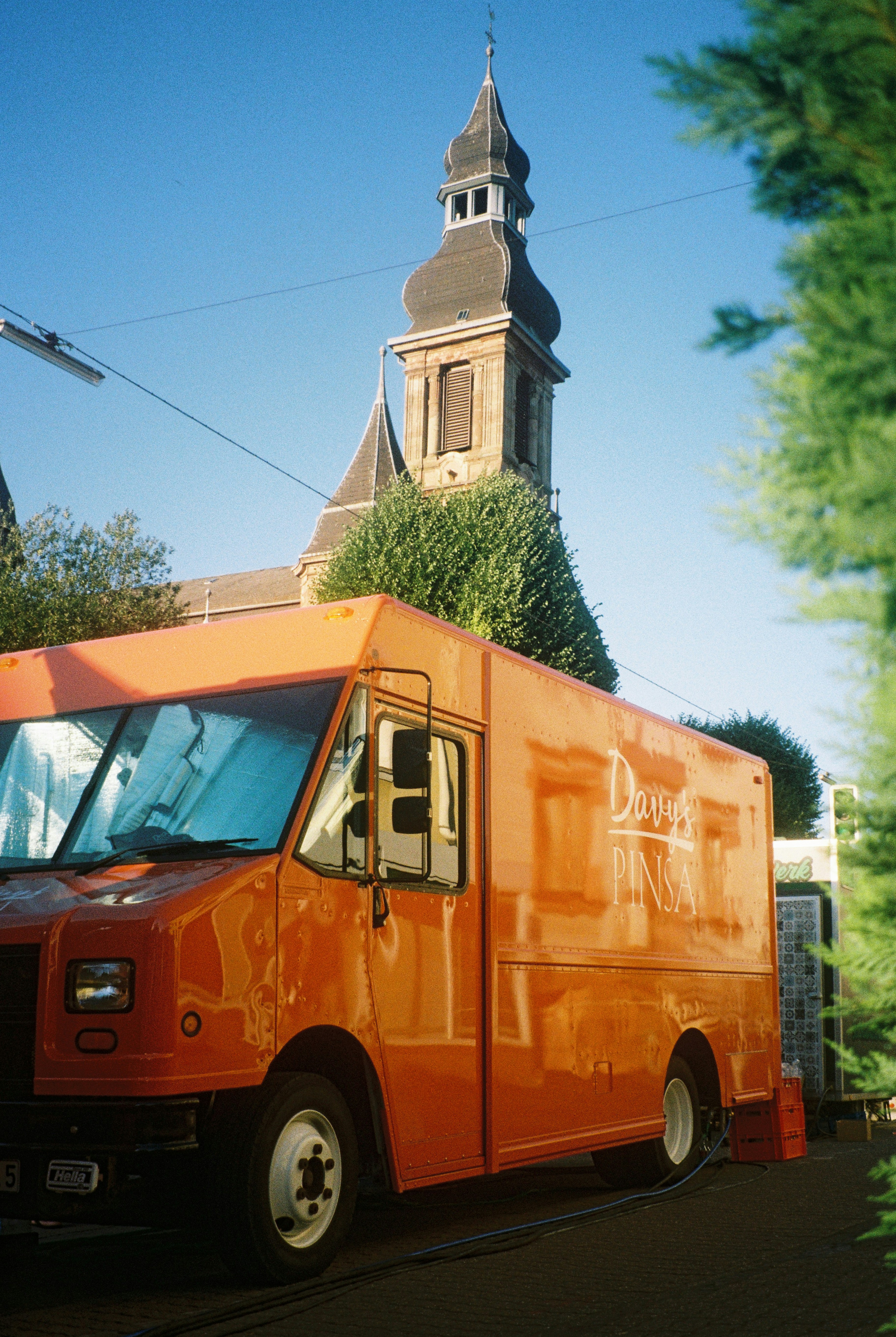 Vibrant orange food truck named 'Dany's Pinsa' parked in an urban setting, with a historic church tower in the background.