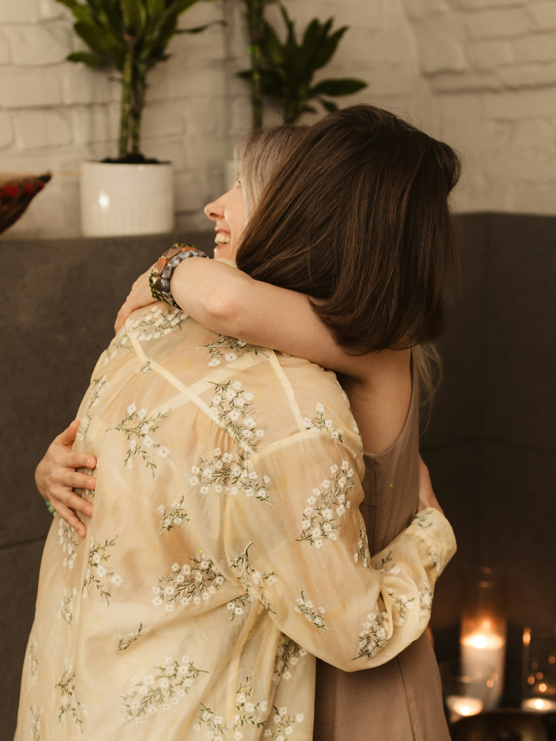 Two individuals share a warm embrace, radiating happiness in a softly lit interior adorned with greenery and candles.