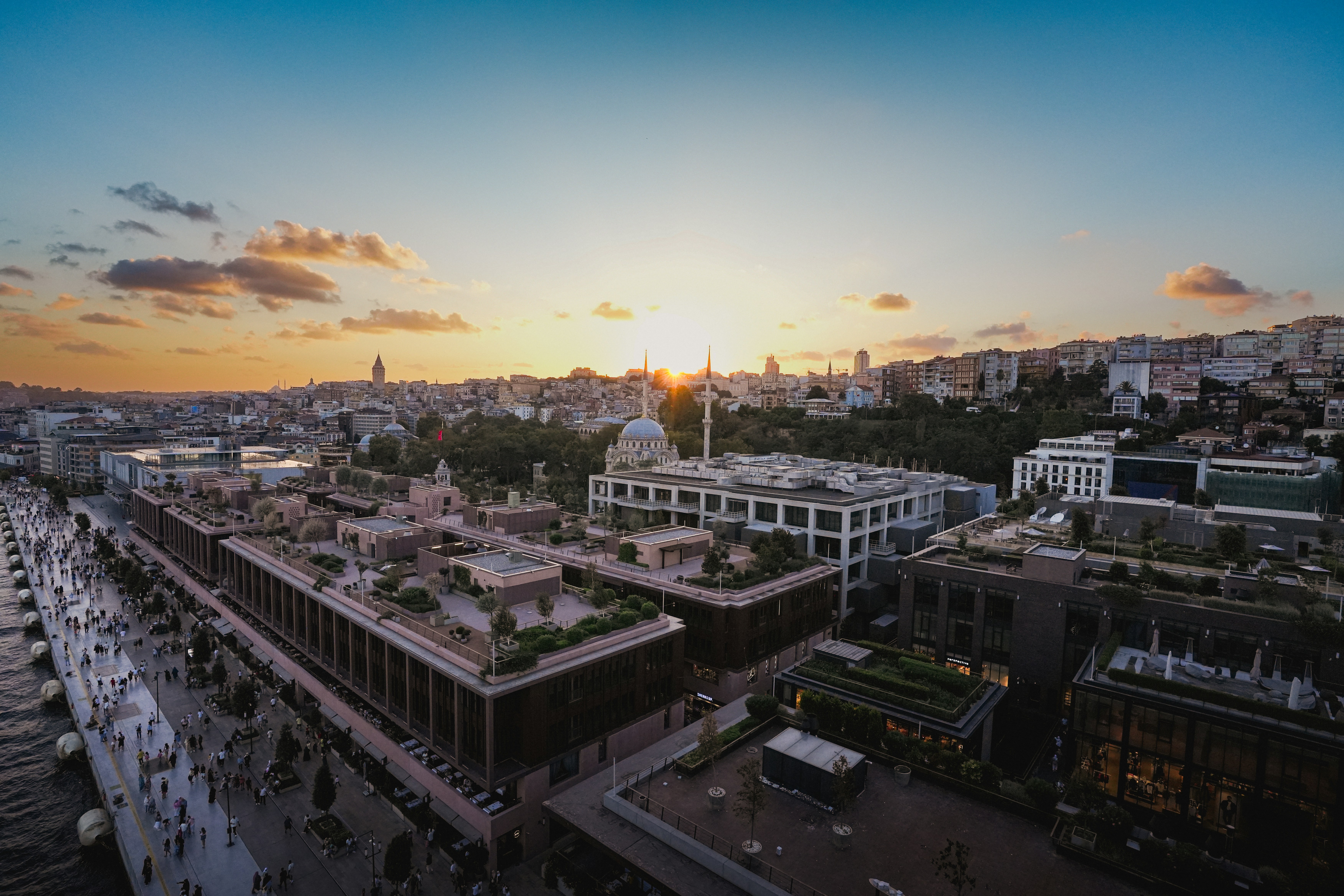 Golden hour in Istanbul — where the sun sets behind the city’s historic skyline, casting a warm glow over the Bosphorus promenade, the Galata Tower in the distance, and the elegant domes and minarets that define this timeless city. | City buildings at sunset with a clear sky.