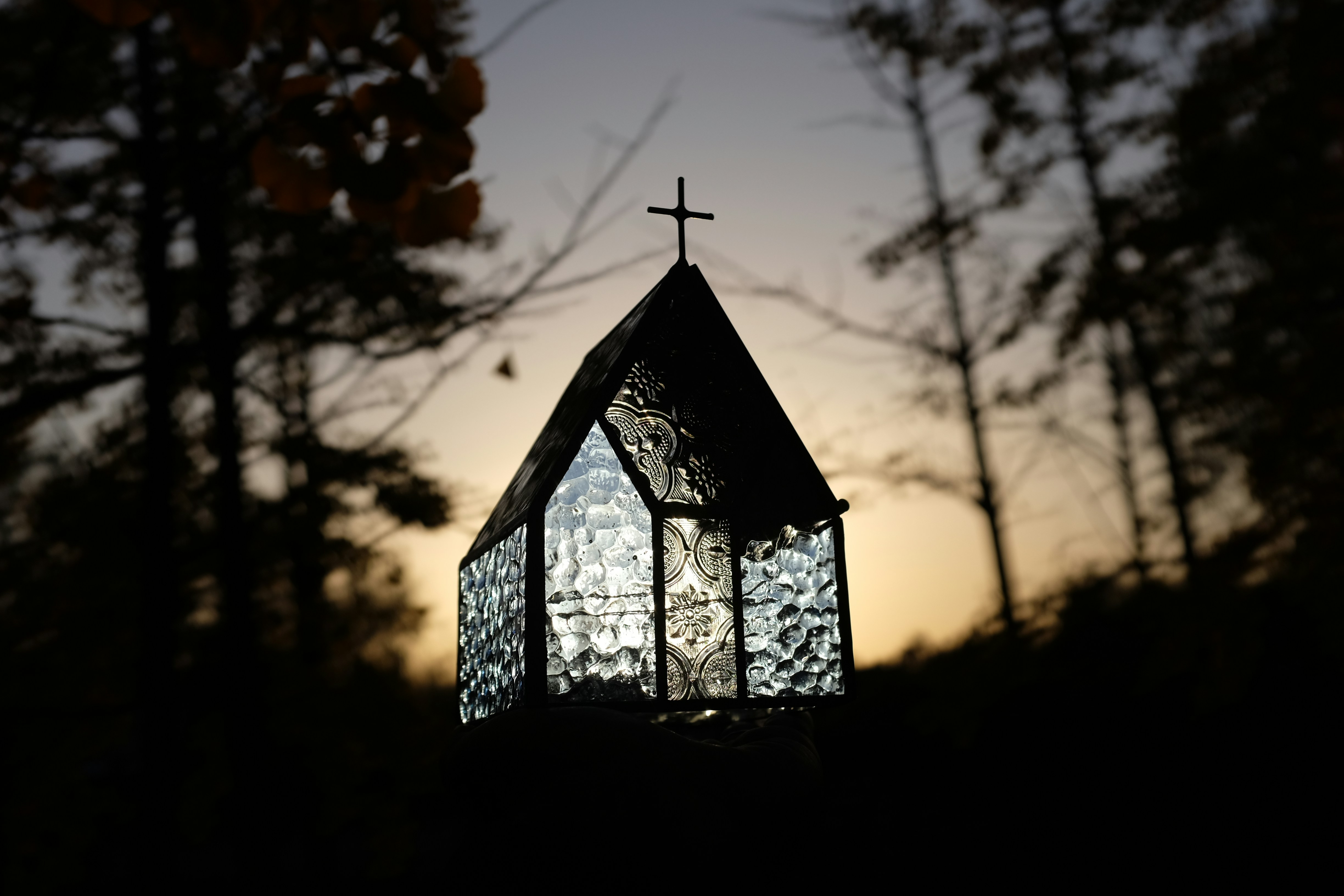 A glass lantern with a cross stands silhouetted against a dusky sky, its textured panels catching faint light; the scene emphasizes quiet spirituality and twilight ambience.