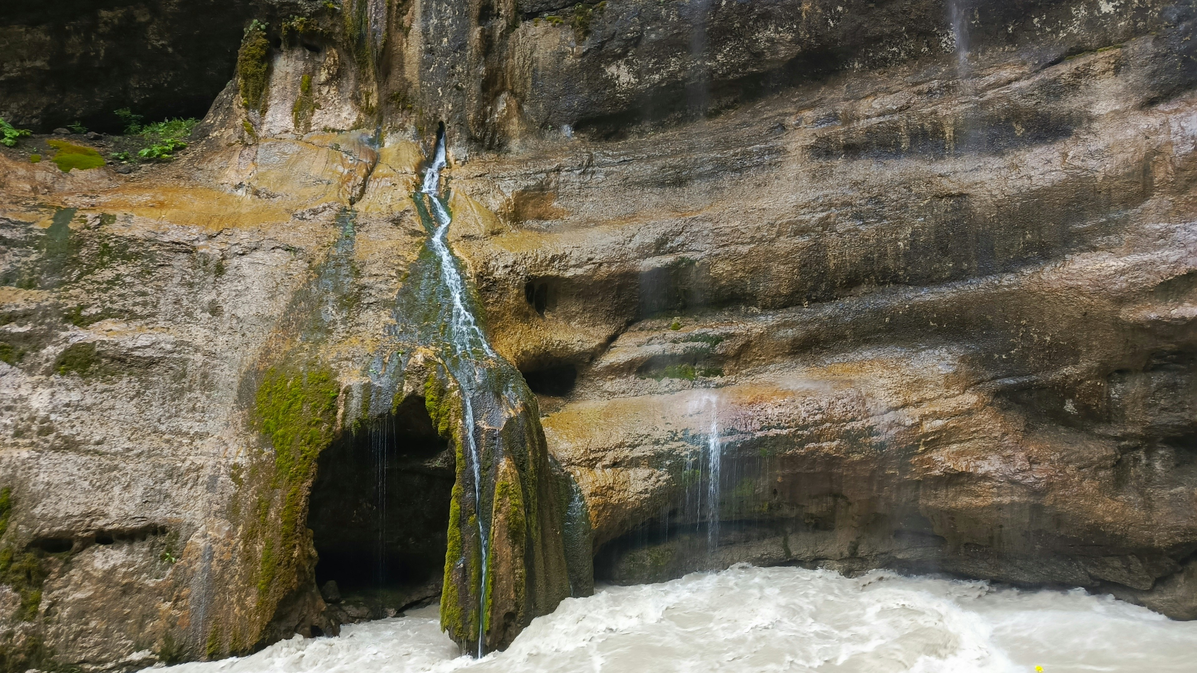 A cascading waterfall trickles down a rugged rock face, surrounded by lush greenery and turbulent waters below.