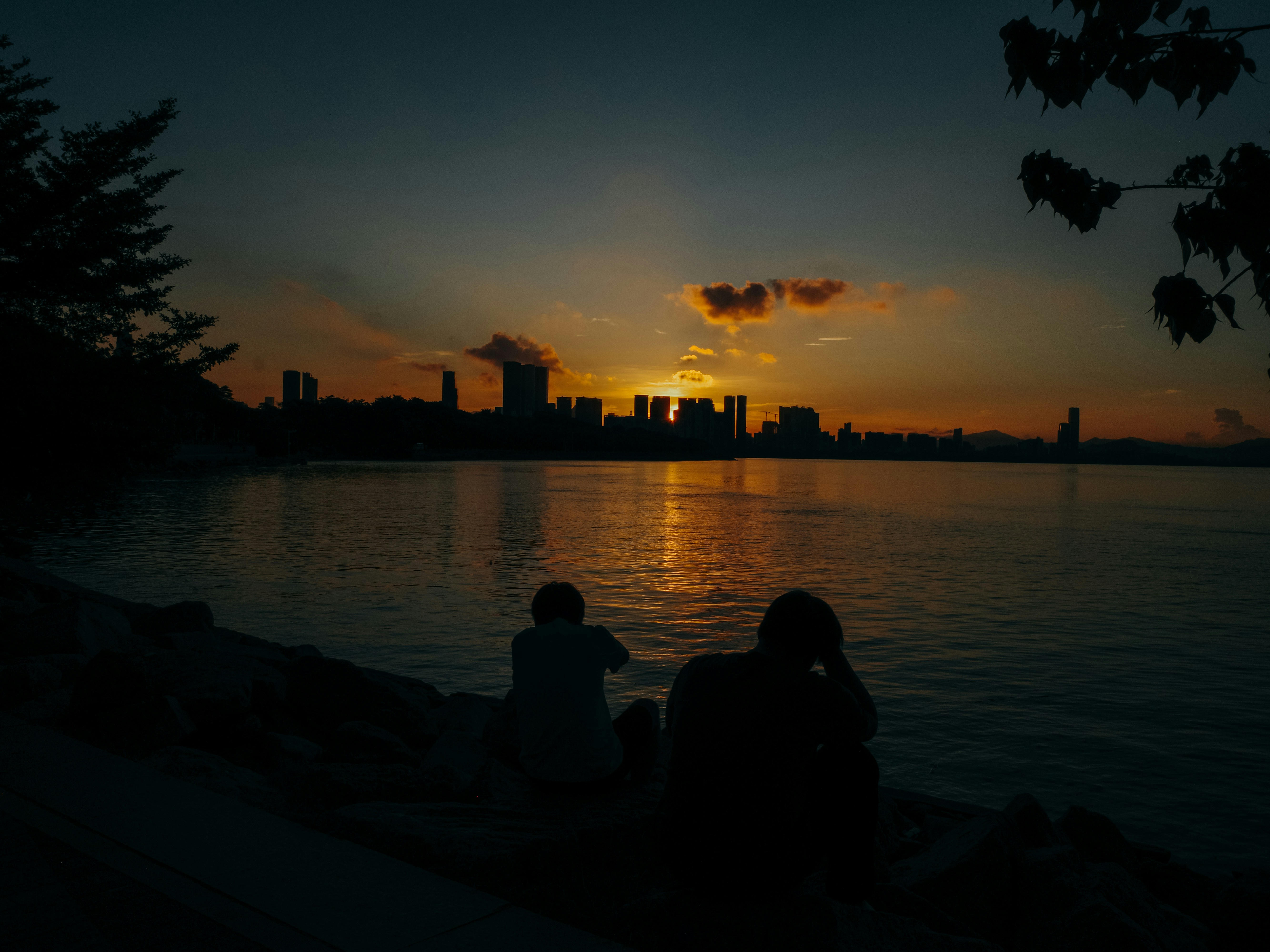 Silhouetted figures sit along the waterfront, watching the sunset reflect off the water, with a city skyline in the background. The scene captures a moment of tranquility as day transitions to night.