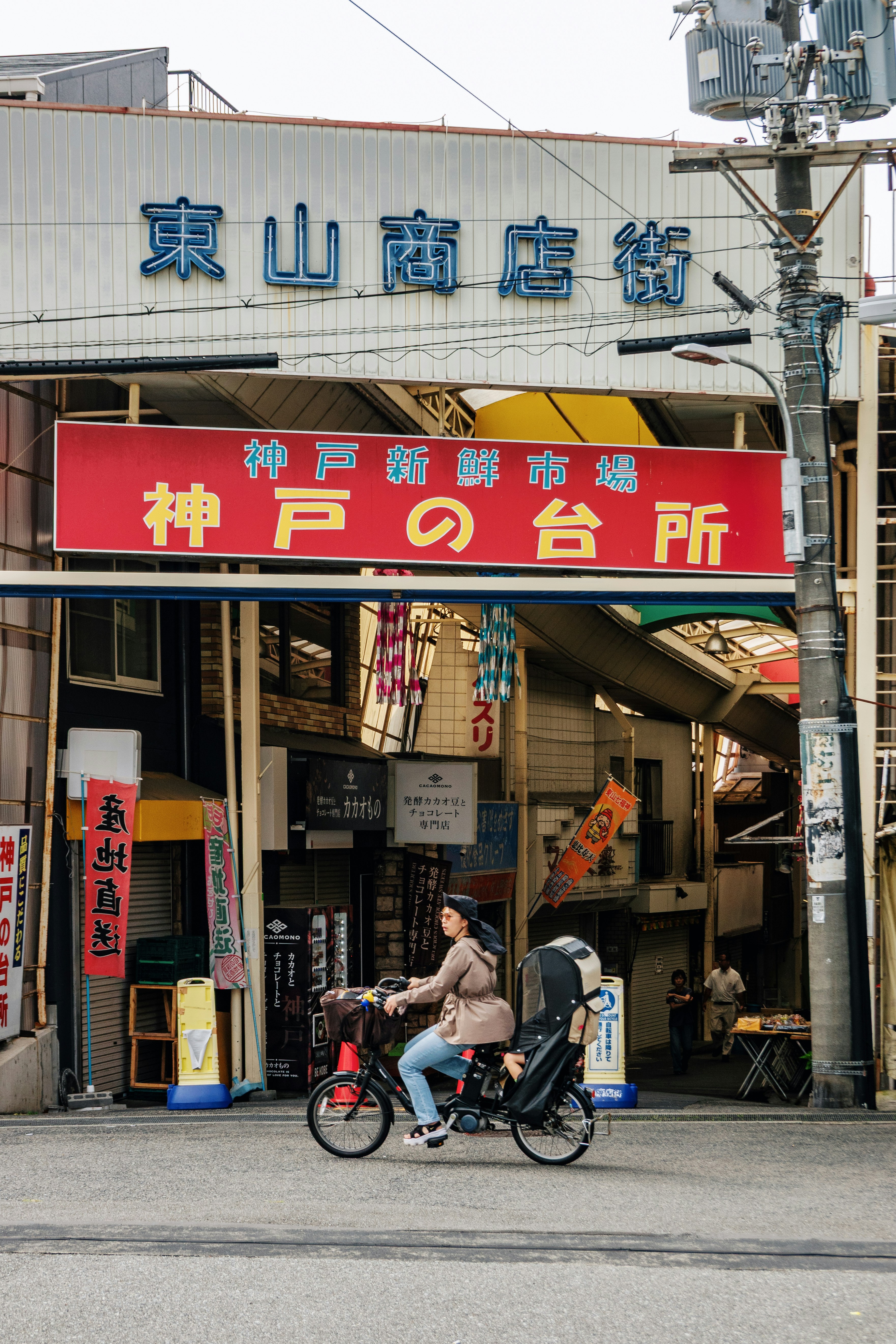 A cyclist navigates through a bustling market entrance adorned with vibrant signage in Kobe, Japan.