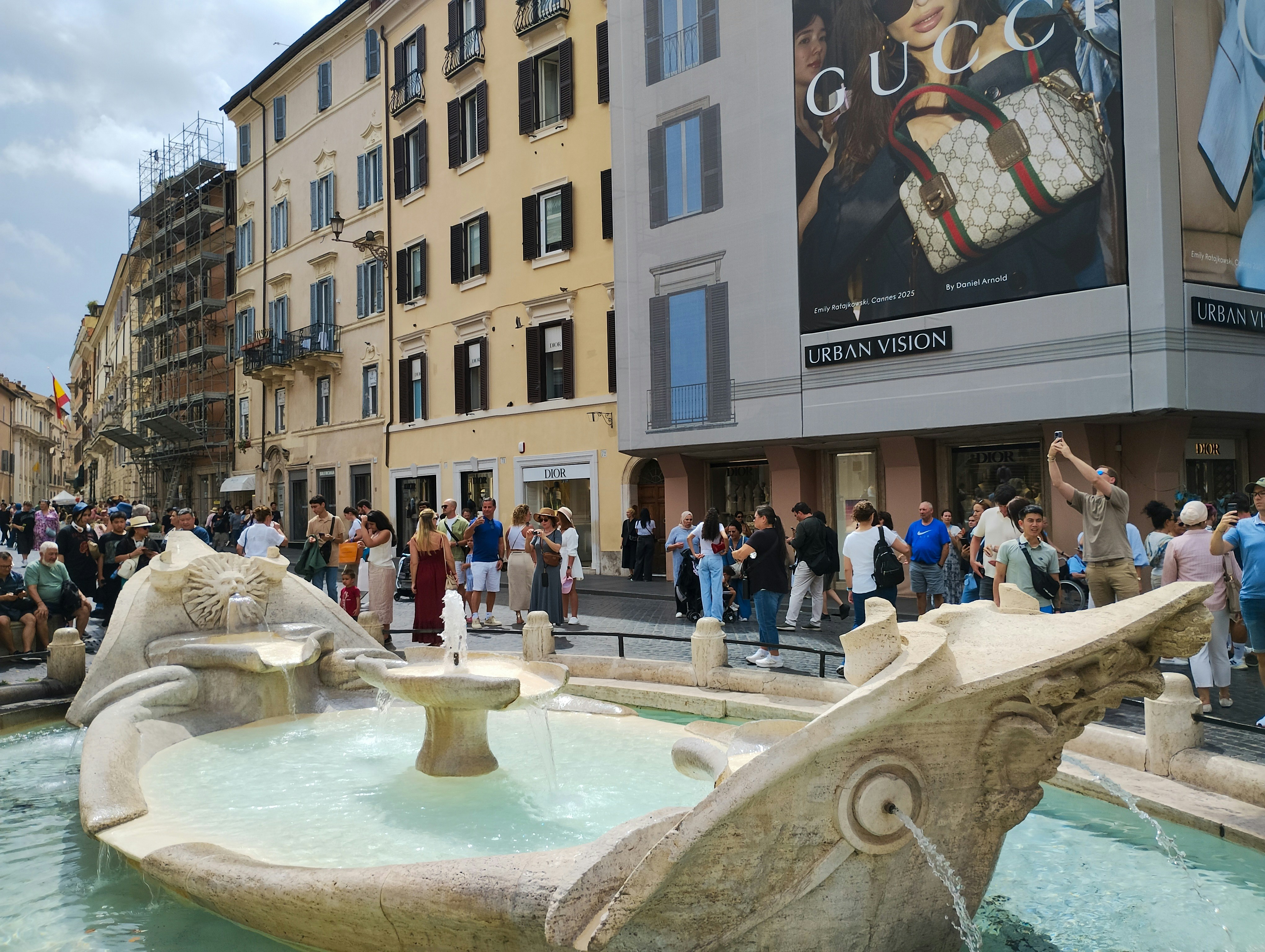 A fountain in a crowded street in Rome, Italy. | Barcaccia fountain with people on a street.
