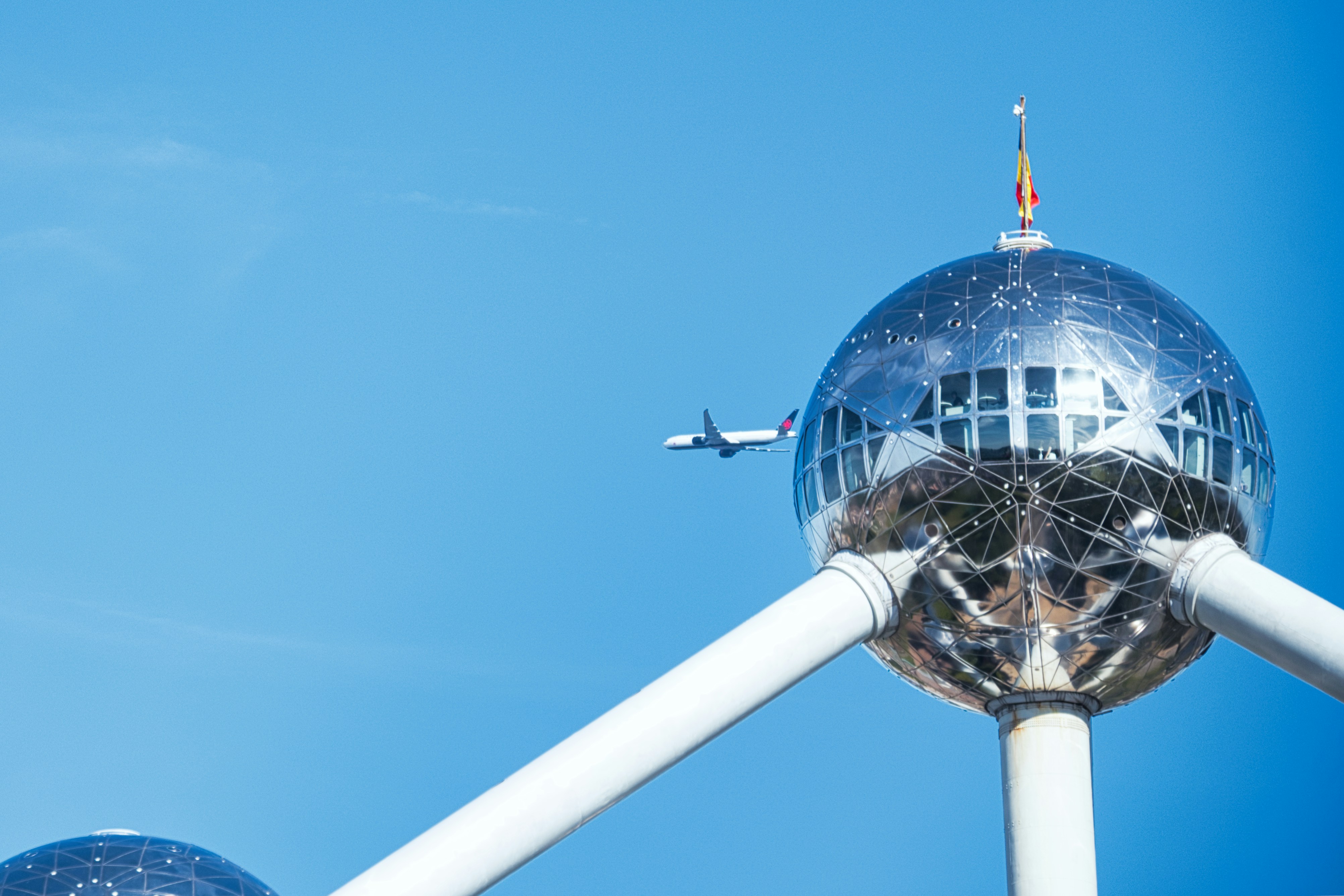 A passenger plane flying over the Atomium in Brussels. | Airplane flying past the atomium structure under a clear blue sky.