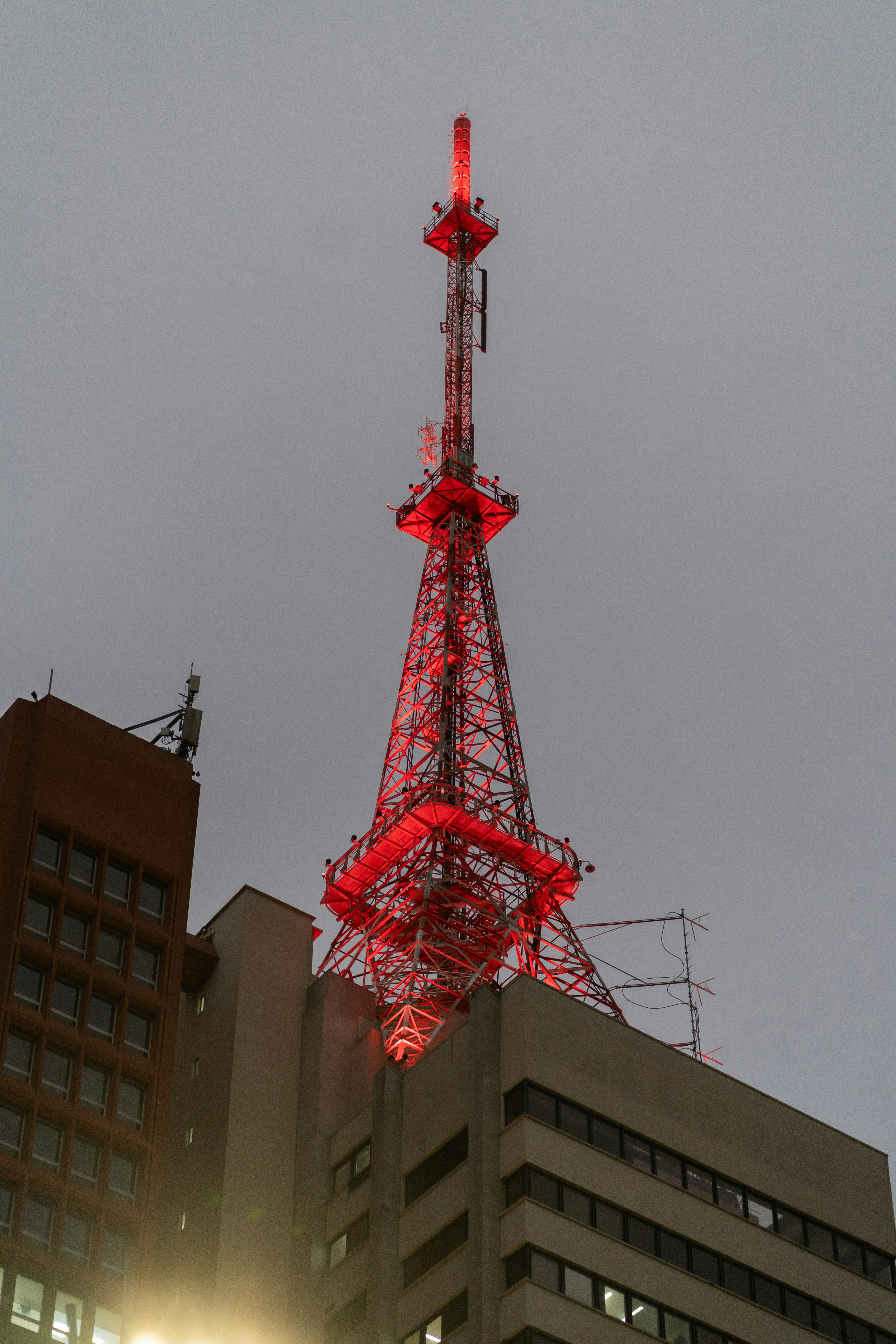 Illuminated communication tower with a striking red glow atop a city building, set against a gray sky.
