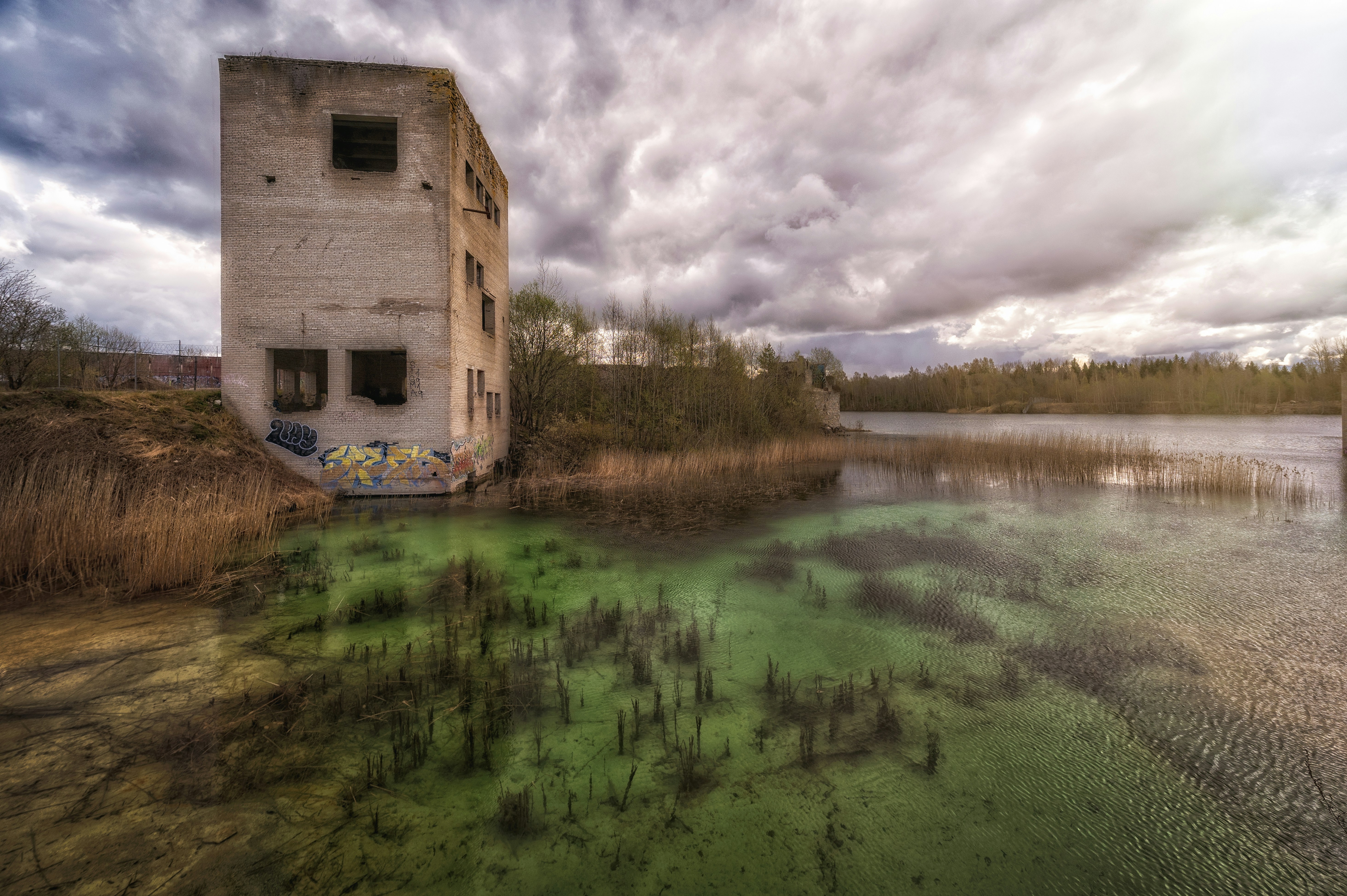 Dilapidated building beside a tranquil, green-tinted lake, surrounded by overgrown reeds and dramatic clouds above. The scene reflects the intersection of nature and urban decay.