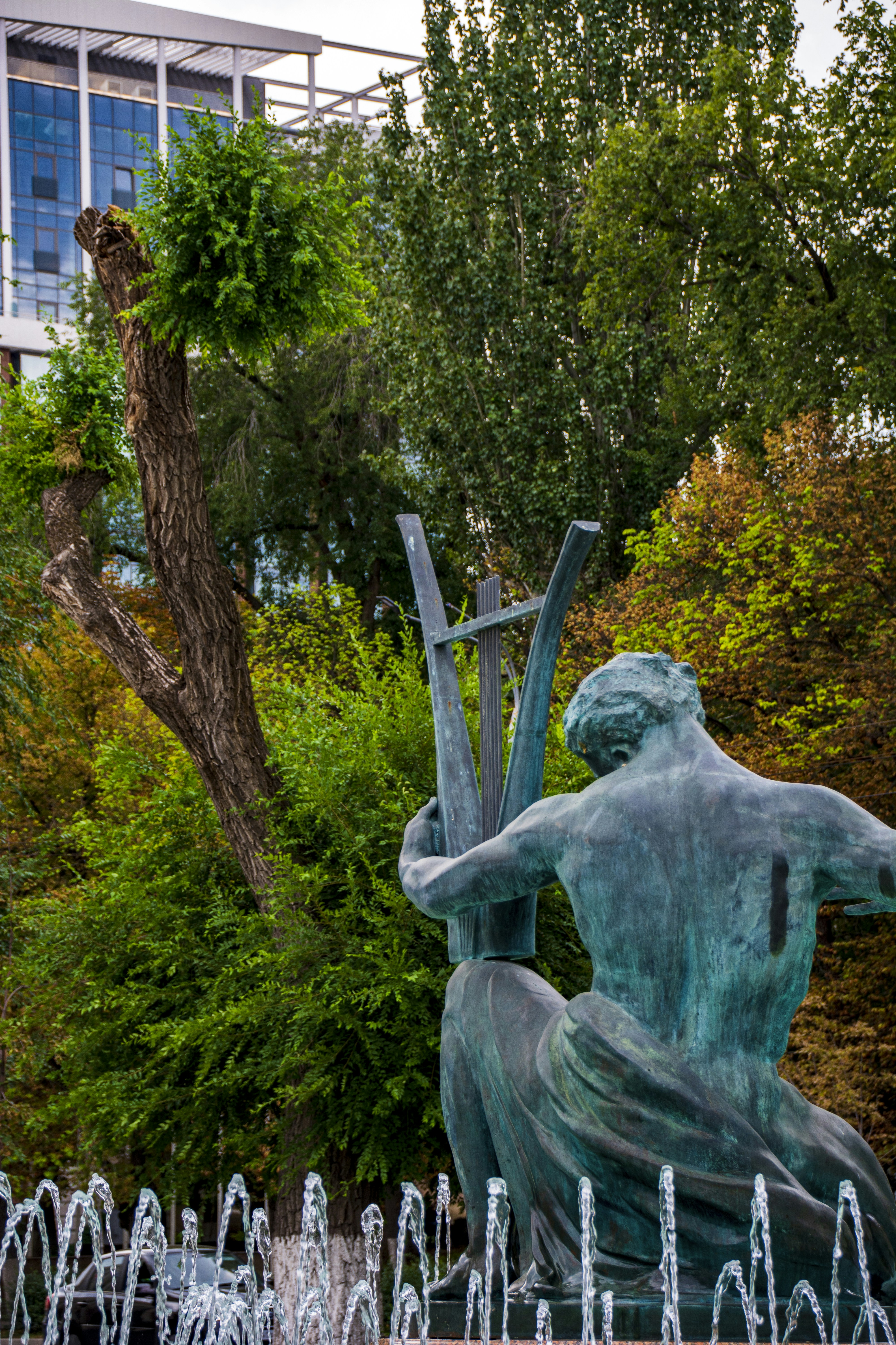 Bronze statue of a musician poised with a harp amidst lush greenery and a fountain. The scene reflects a serene blend of art and nature.
