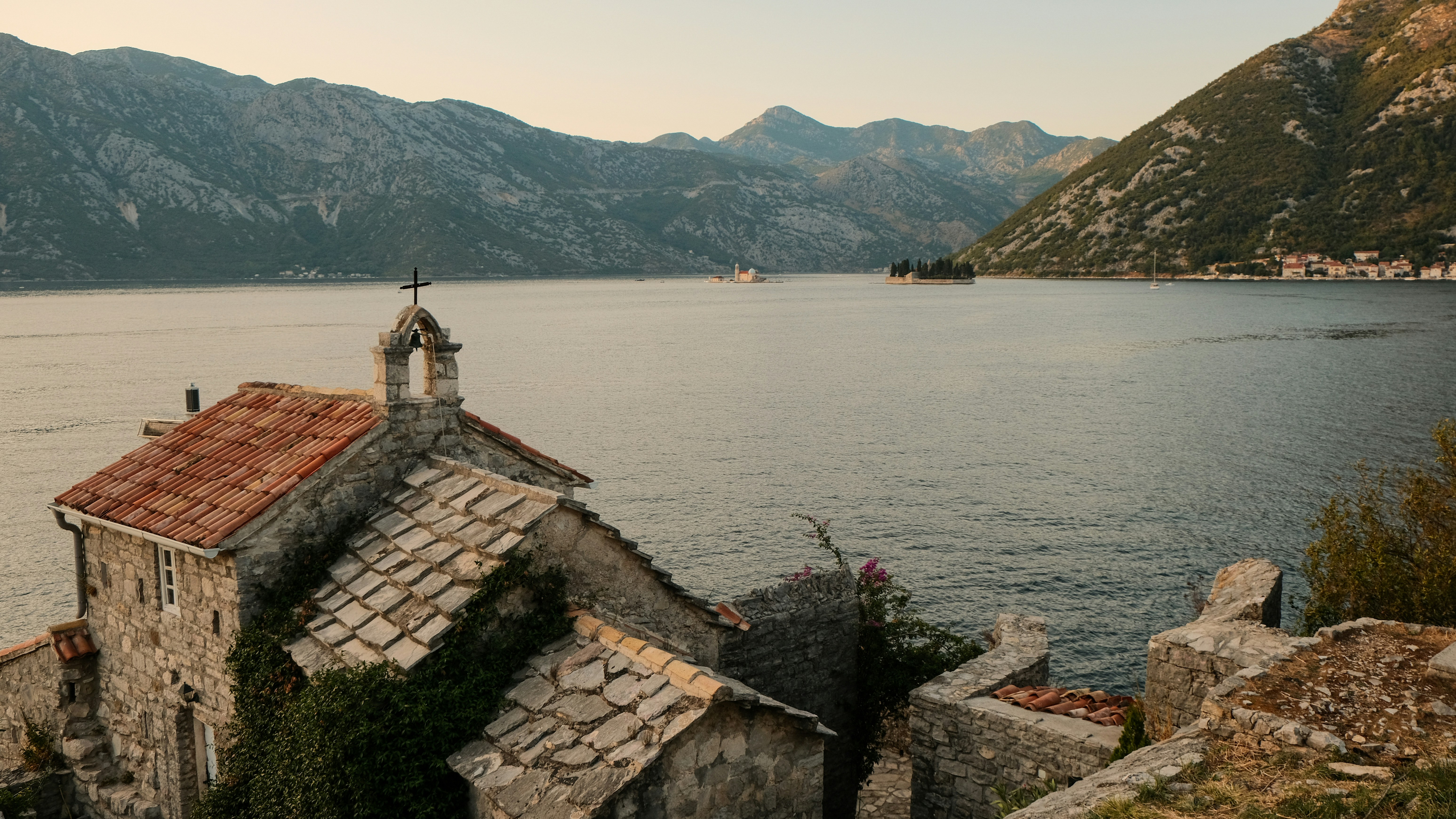 Historic stone house adorned with creeping vines overlooks a tranquil bay, framed by majestic mountains. The scene captures the essence of serene coastal living.