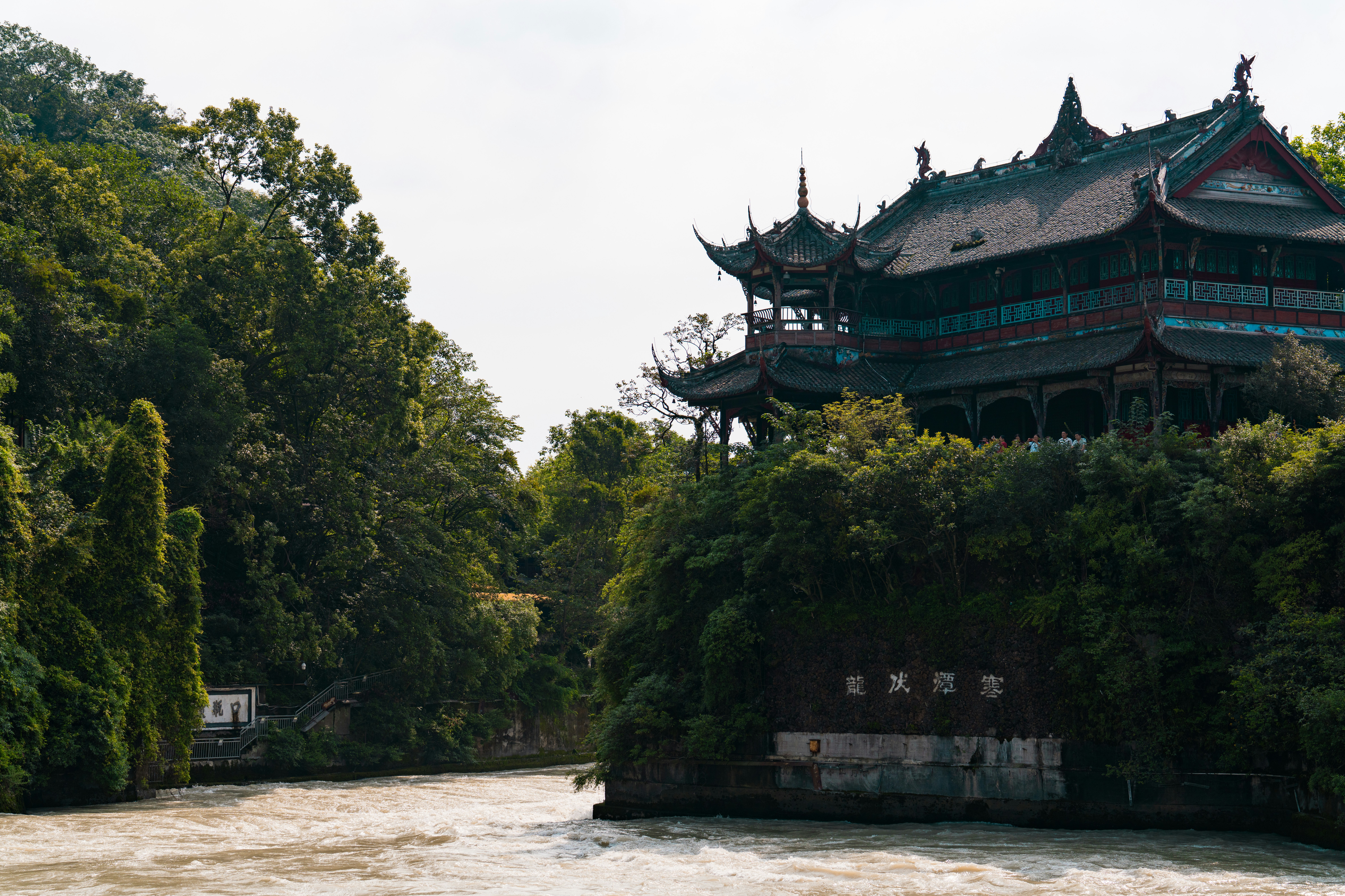 Traditional Chinese architecture nestled among lush greenery, overlooking a flowing river.