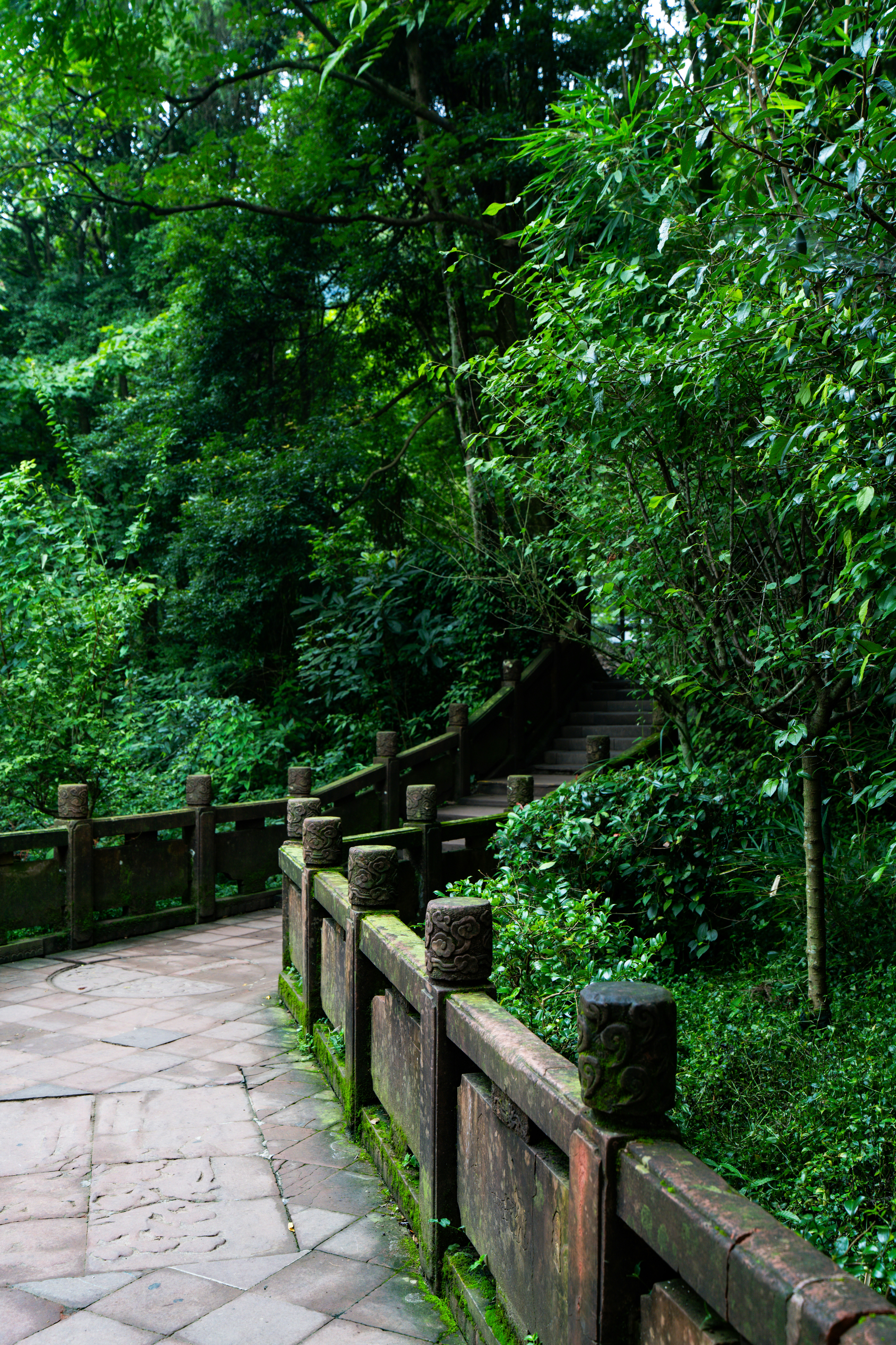 Winding stone path bordered by intricately carved wooden railings, surrounded by lush greenery in a serene forest setting.