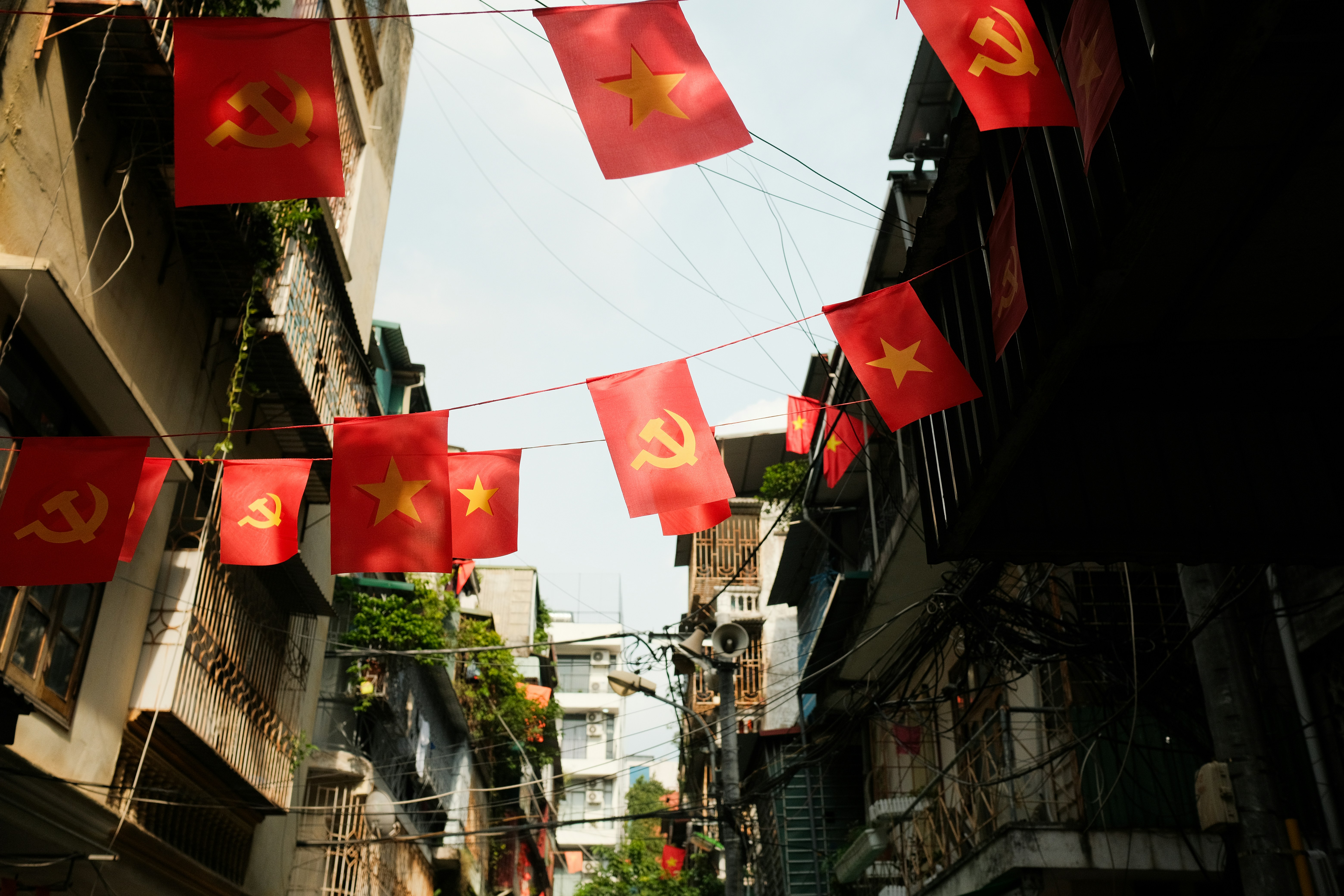 Vibrant red flags featuring the hammer and sickle hang above a narrow alley, surrounded by urban architecture. The scene captures a moment of cultural significance.
