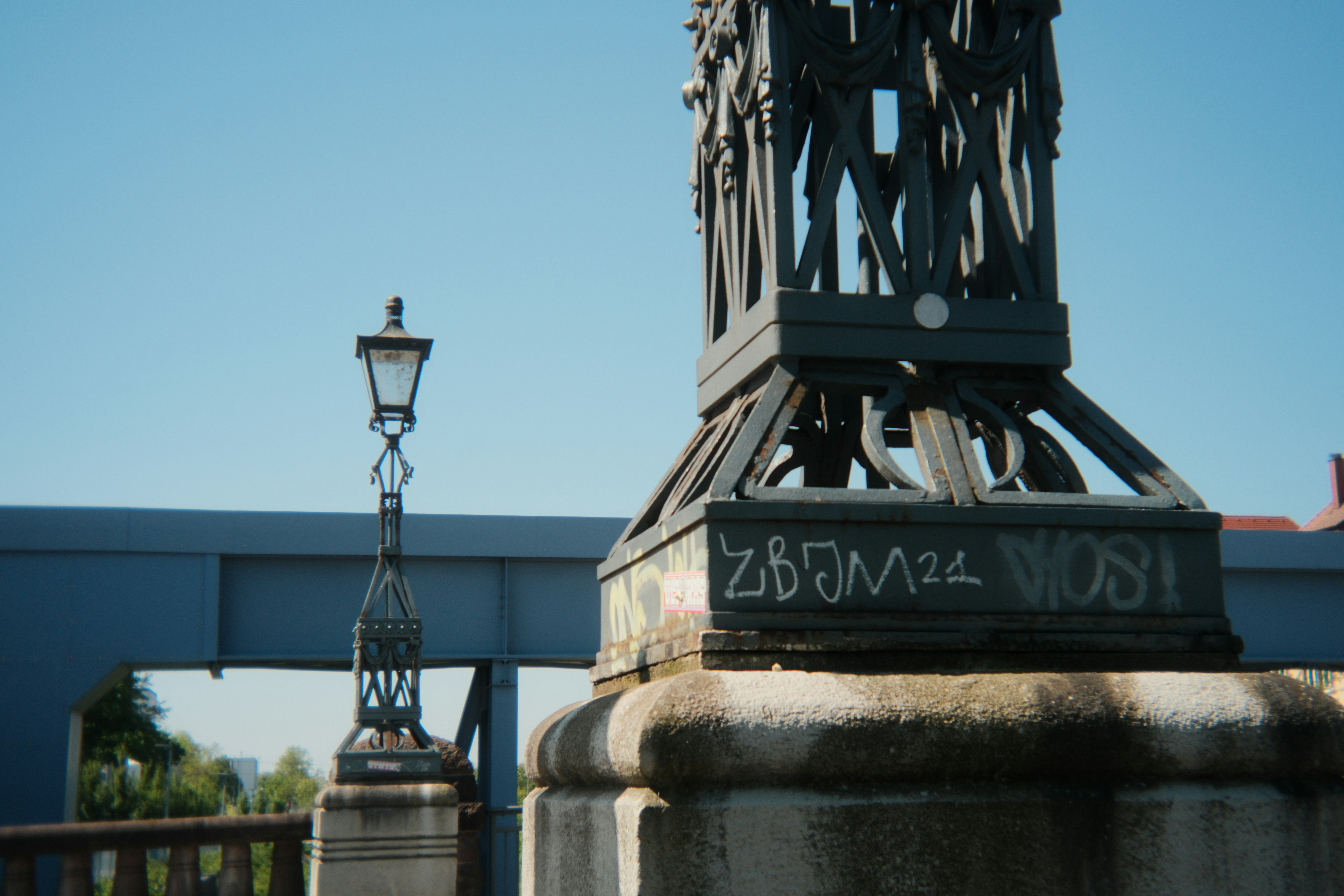 Intricate metalwork of a lamp post juxtaposed against graffiti on a stone base, framed by a clear blue sky. The scene captures a moment of urban artistry.