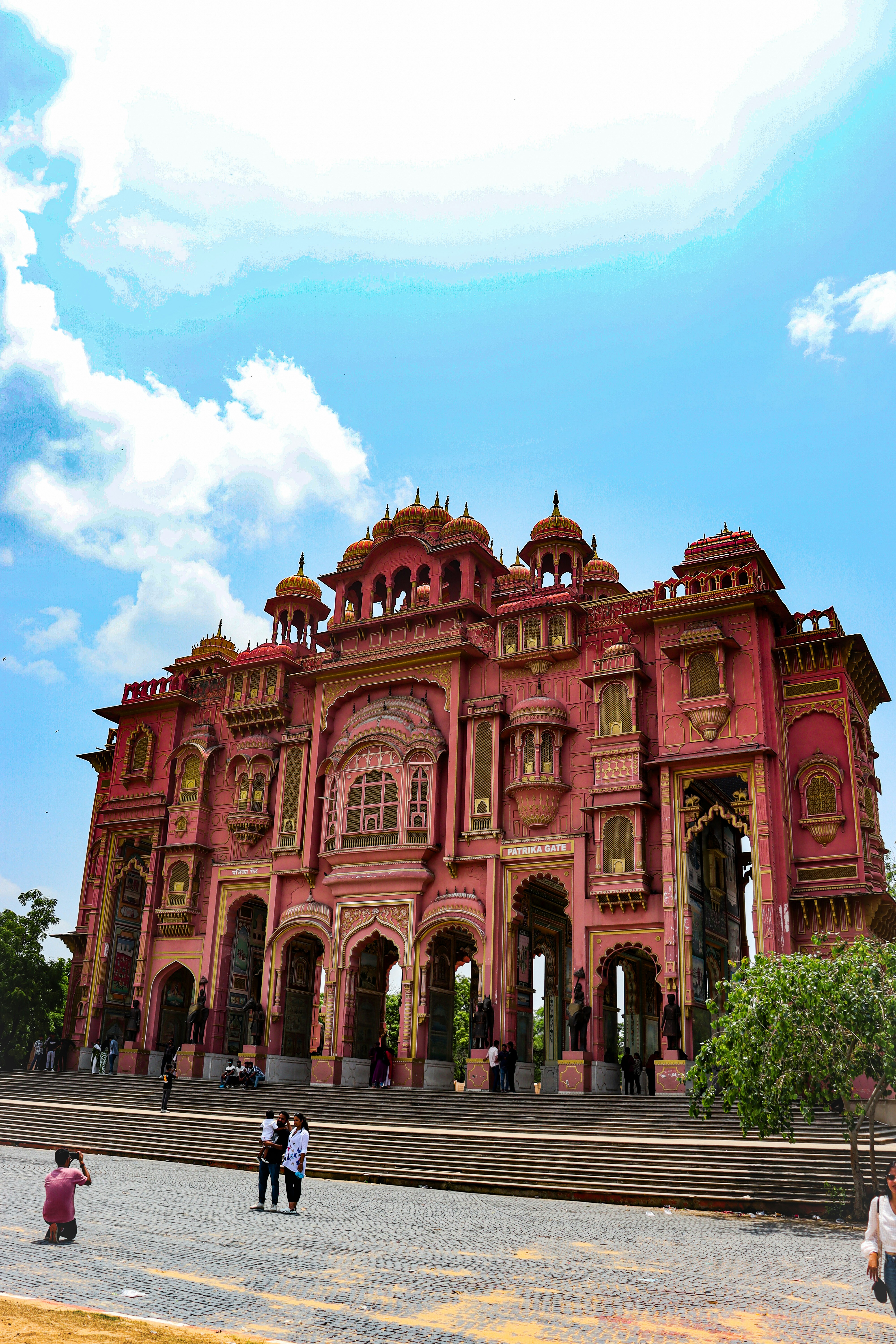 The ornate pink Patrika Gate in Jaipur, Rajasthan, stands grandly under a bright blue sky as visitors gather on the steps. | Ornate pink palace entrance with intricate architectural details