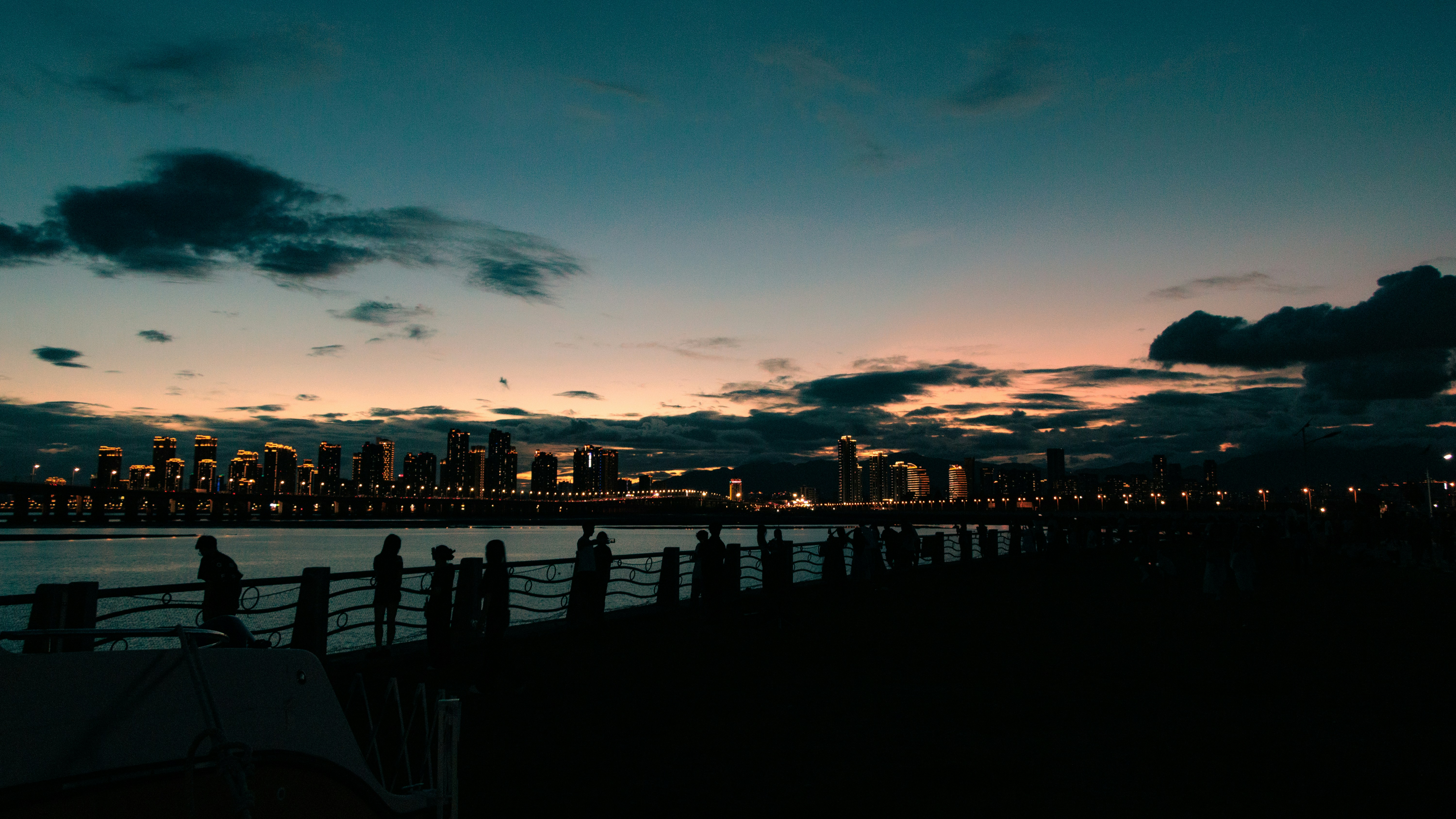Silhouettes of people along a waterfront promenade as city lights twinkle against a twilight sky. The scene captures the serene transition from day to night.