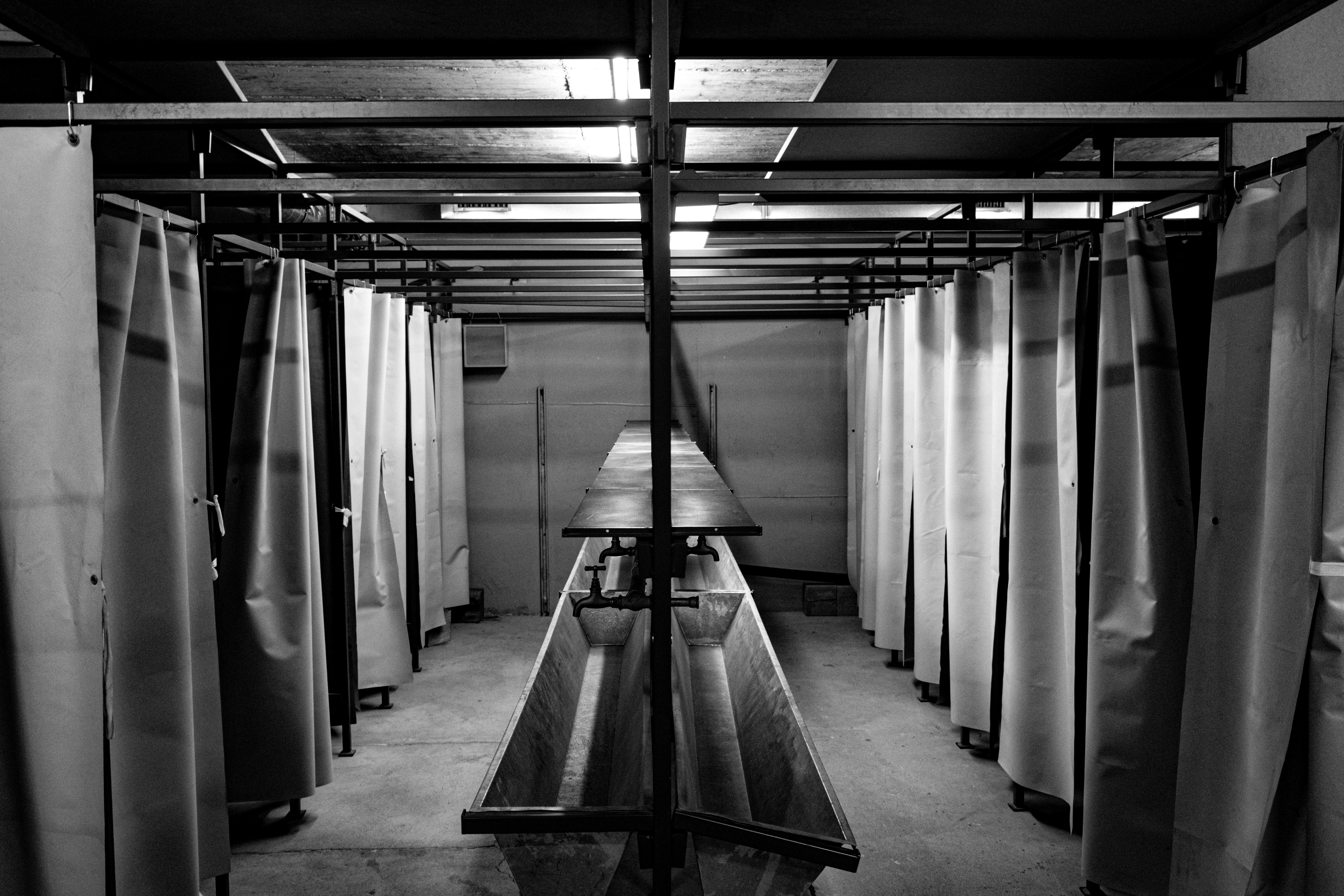 Cold War washing facilities inside the Sonnenberg bunker in Lucerne, Switzerland. Black and white photo of communal wash basins and shower stalls with curtains, reflecting the stark functionality of underground civil defense shelters. | Rows of welding booths with curtains in a workshop.
