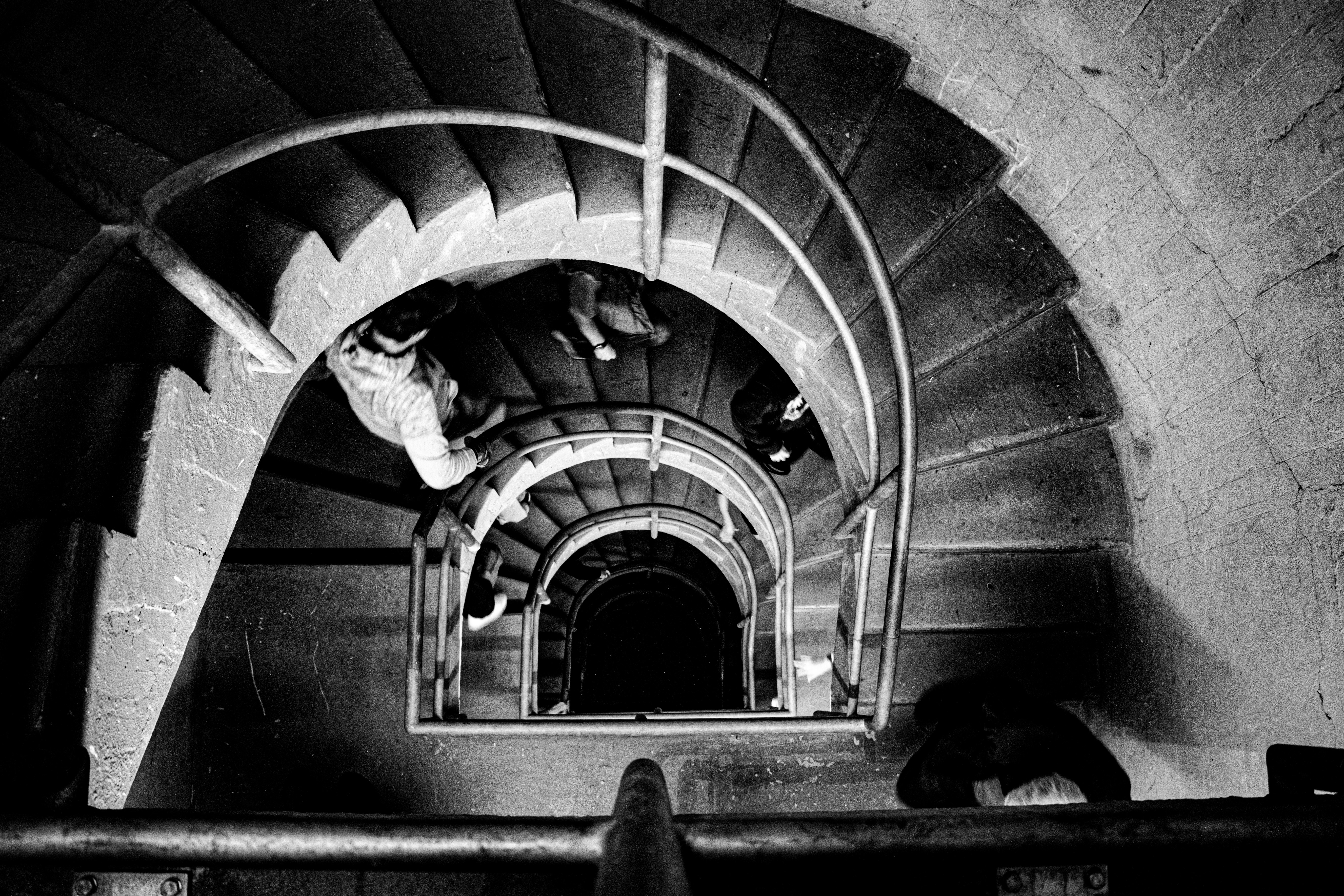 Cold War staircase inside the Sonnenberg bunker in Lucerne, Switzerland. Black and white photo of a spiral concrete staircase with people descending, symbolizing the massive underground scale of civil defense infrastructure. | People descending a spiral staircase in a dark tower.