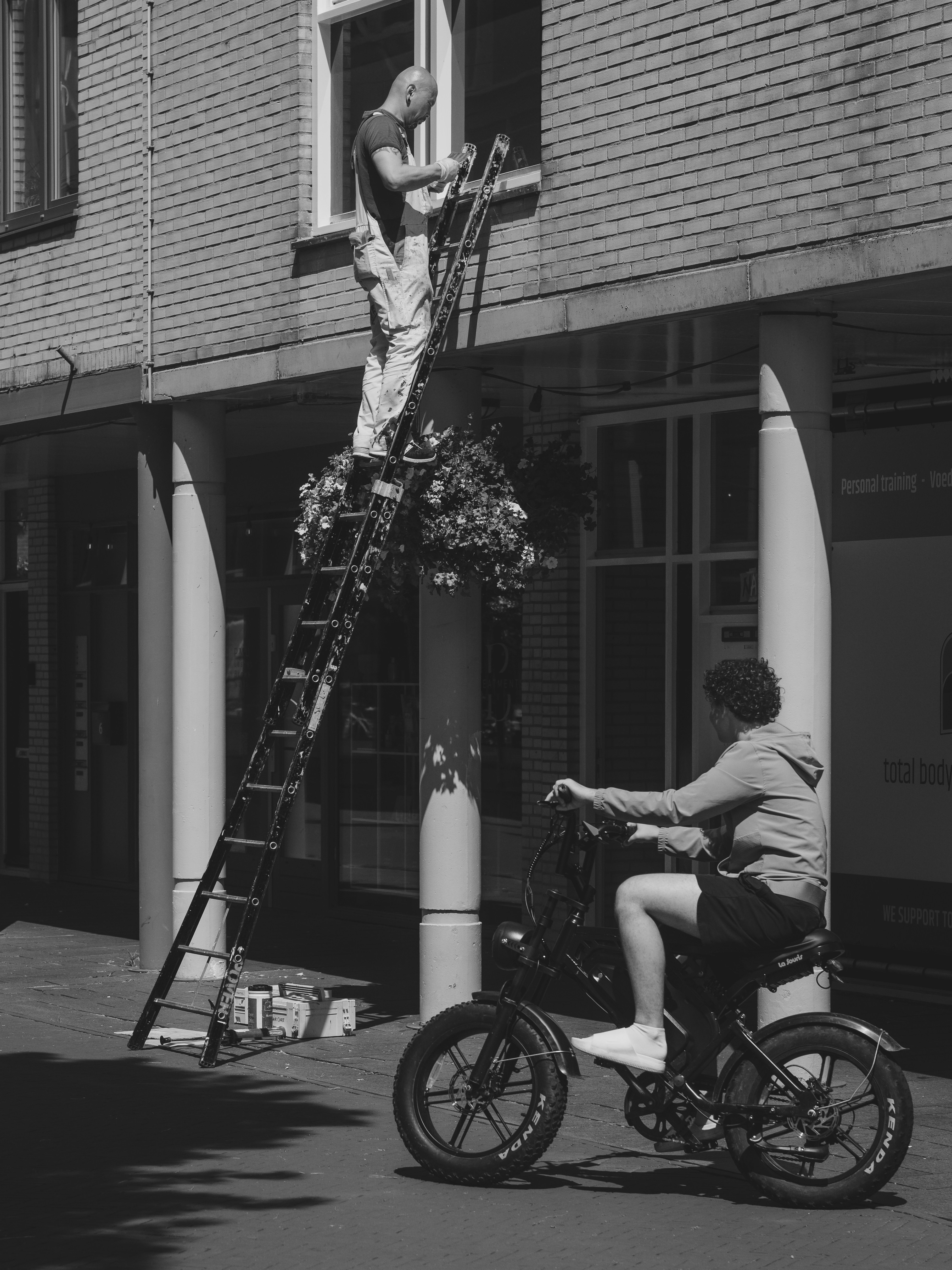 A worker on a ladder attends to a building while a cyclist passes by, capturing the balance of urban life. The scene is depicted in monochrome.