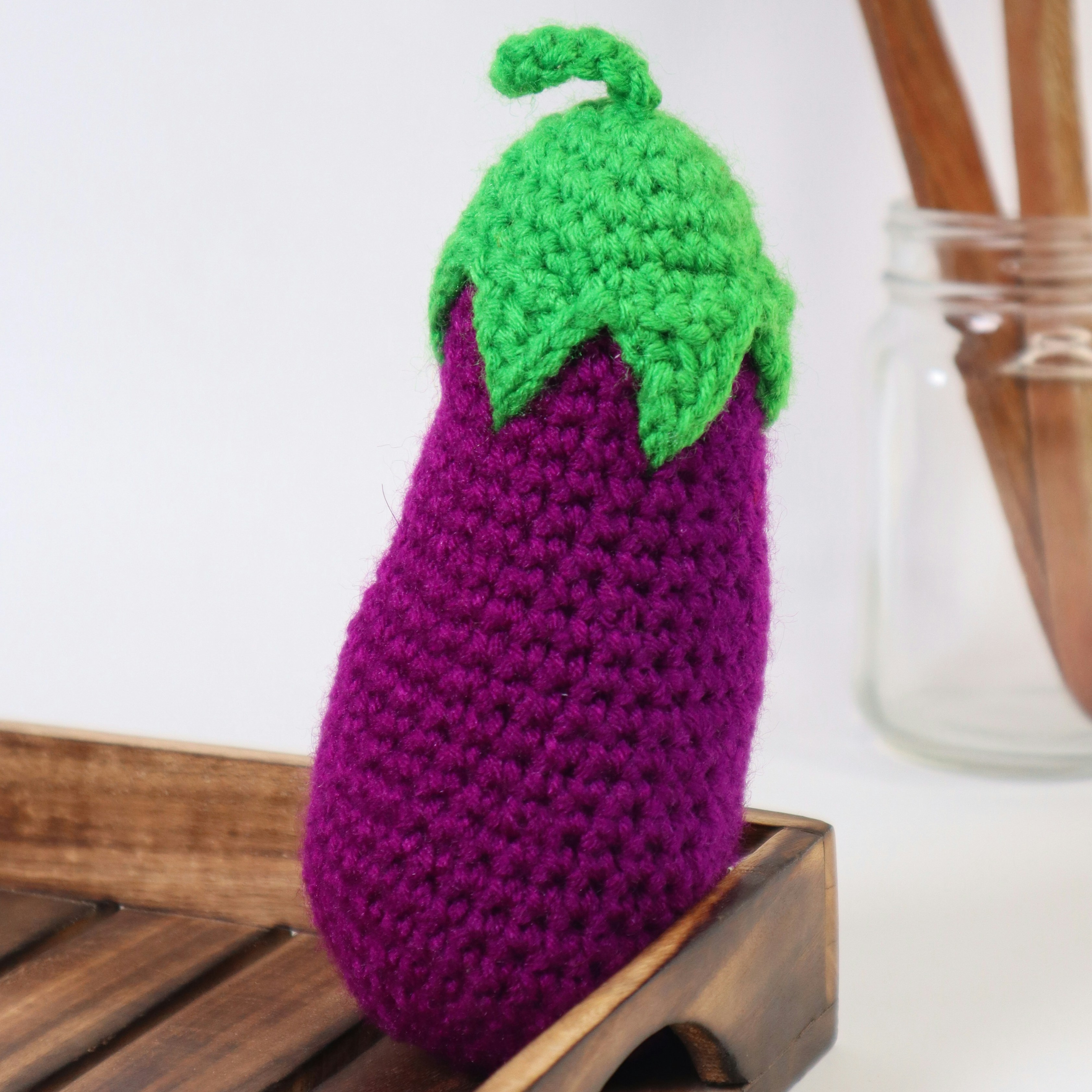 Crocheted eggplant with a vibrant purple body and green top, resting on a wooden surface. A playful representation of a vegetable.