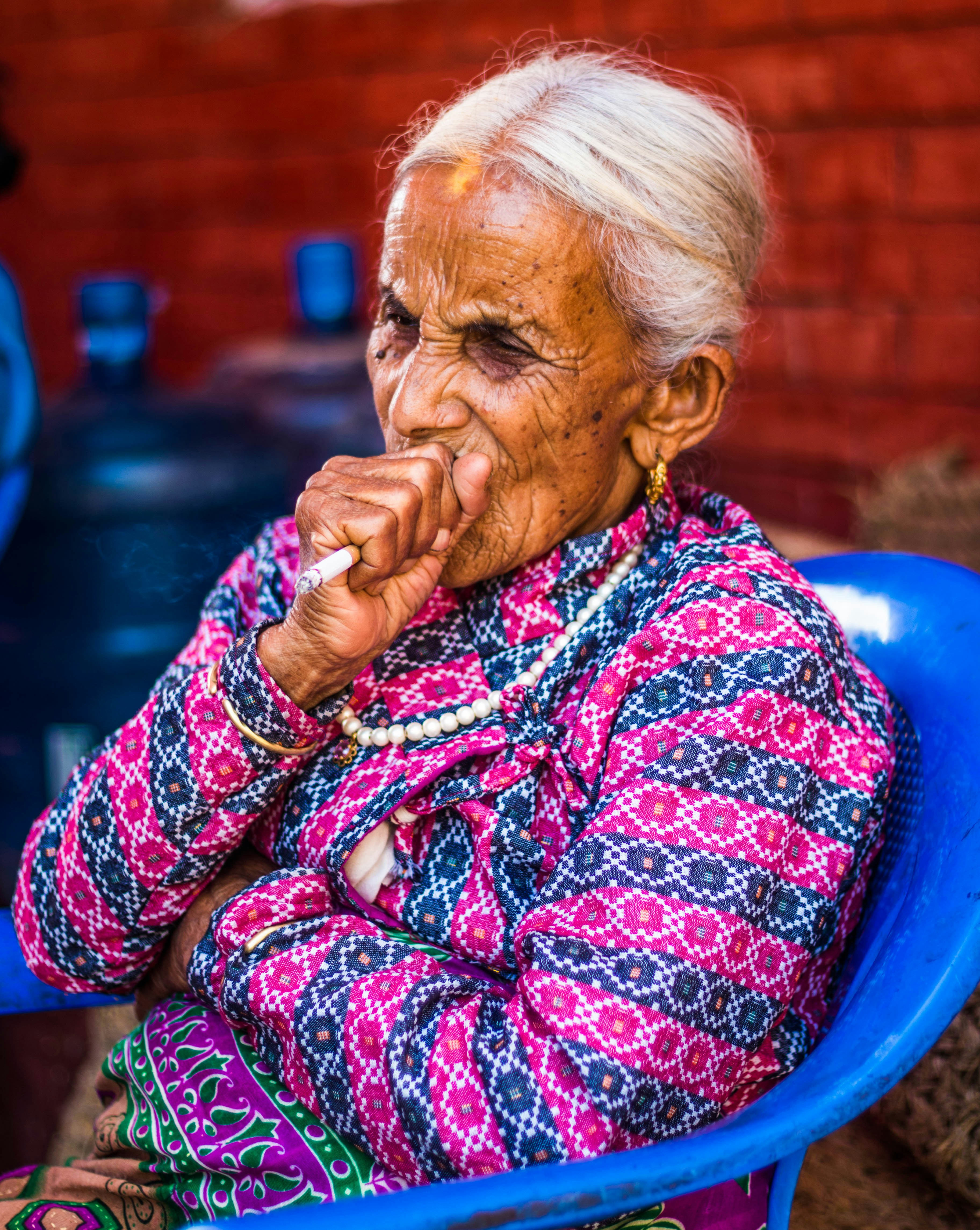 Elderly woman in vibrant traditional attire, lost in thought while holding a cigarette. Background features a rustic setting with colorful elements.