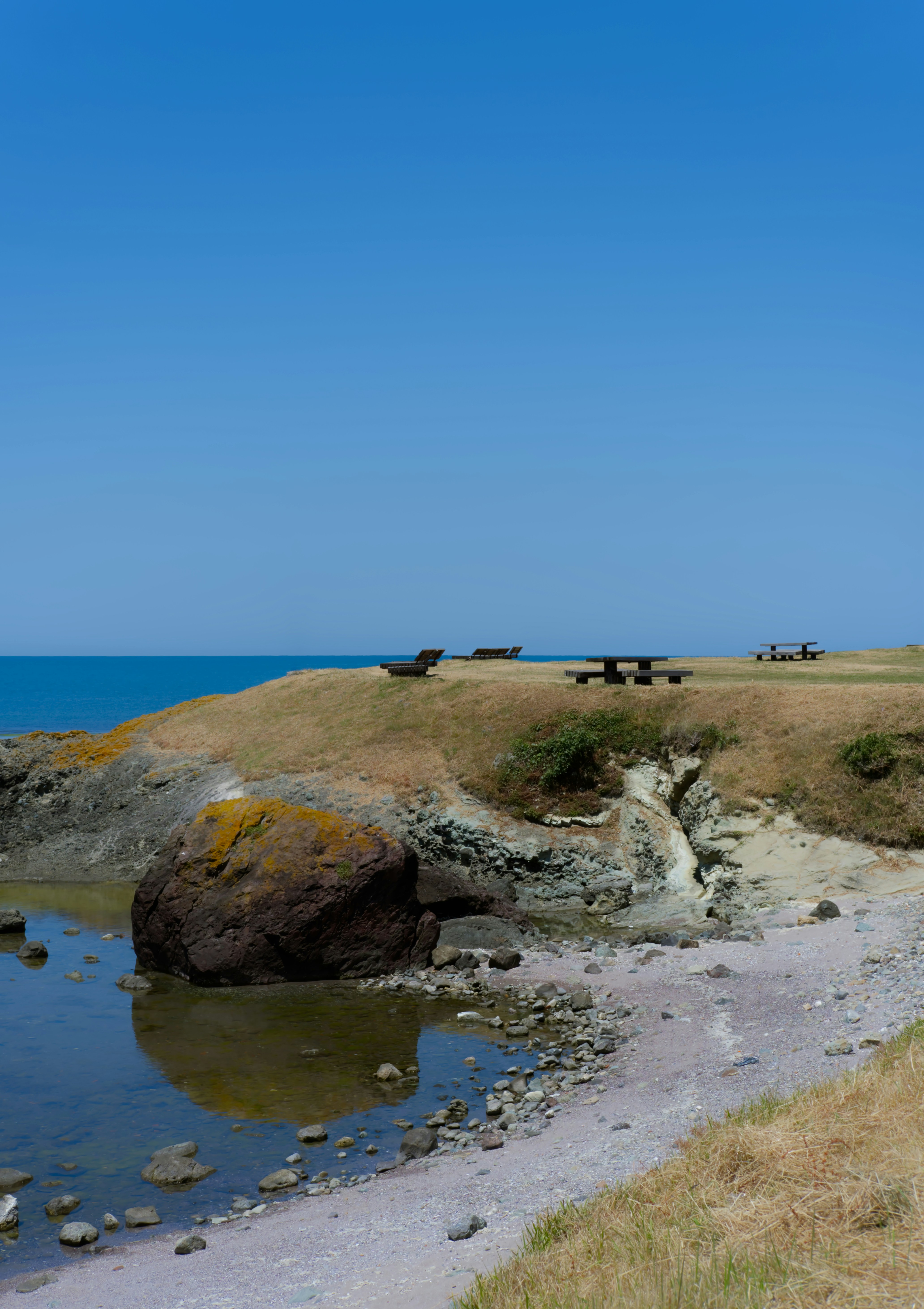 A tranquil coastal scene featuring a rocky shoreline and picnic tables overlooking the calm sea. The clear blue sky complements the peaceful atmosphere.