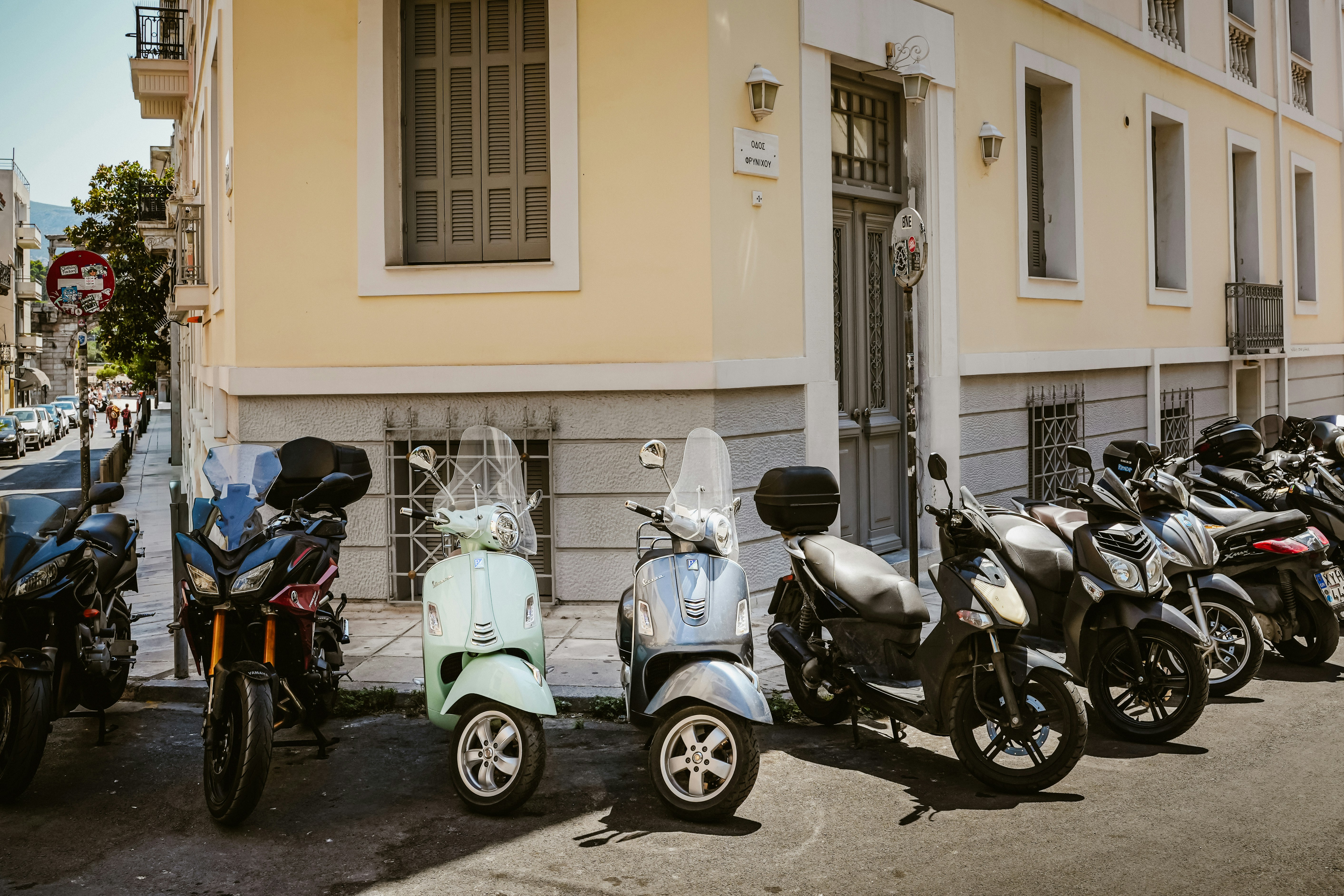 Scooters parked along a residential street in Athens’ Plaka district, in front of a neoclassical-style building with pale yellow walls and shuttered windows. | Several parked scooters and motorcycles on a sunny street.
