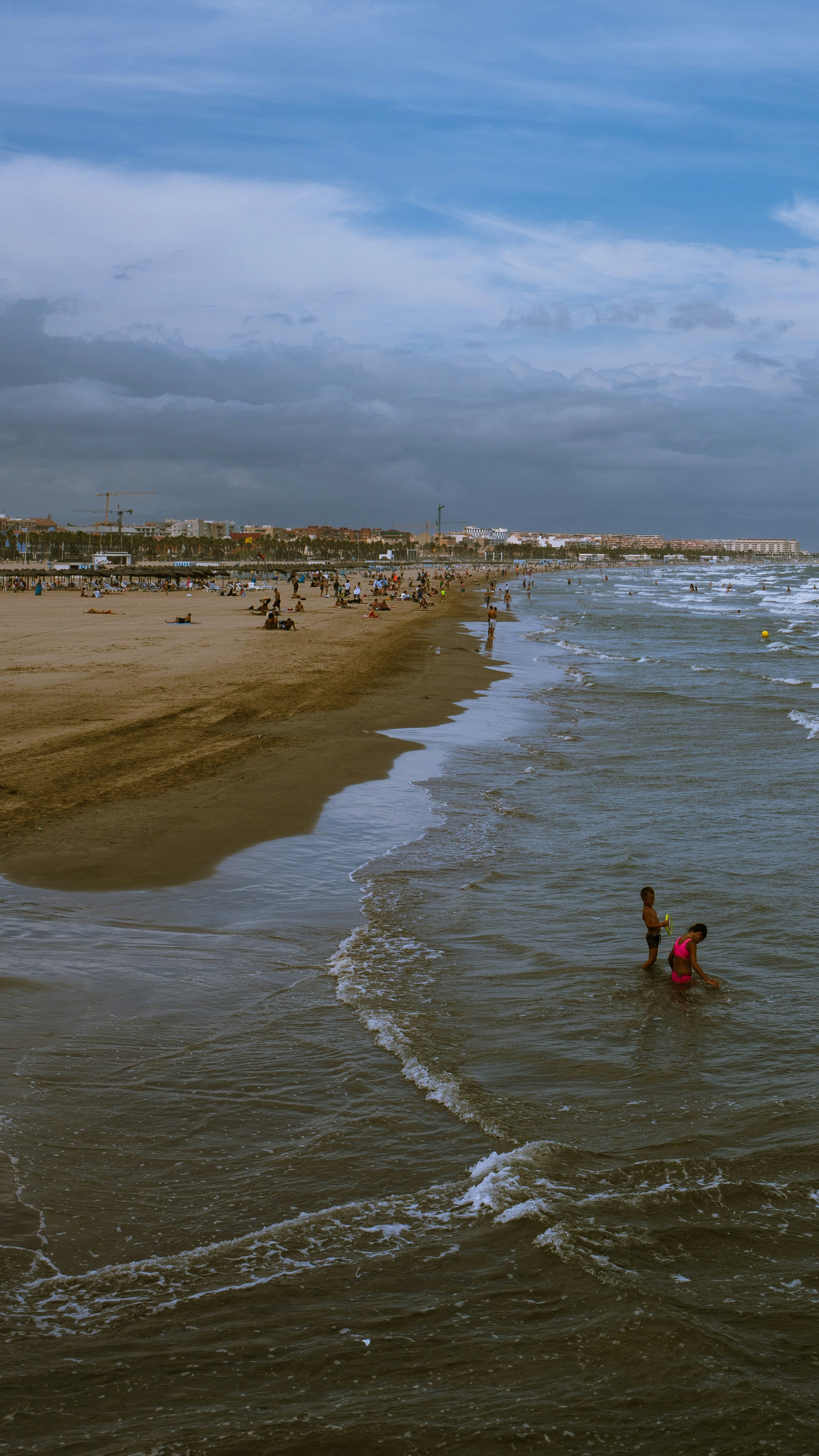 Children play in shallow waves as beachgoers enjoy a sunny day along the coast. The horizon blends with a cloudy sky, creating a serene atmosphere.