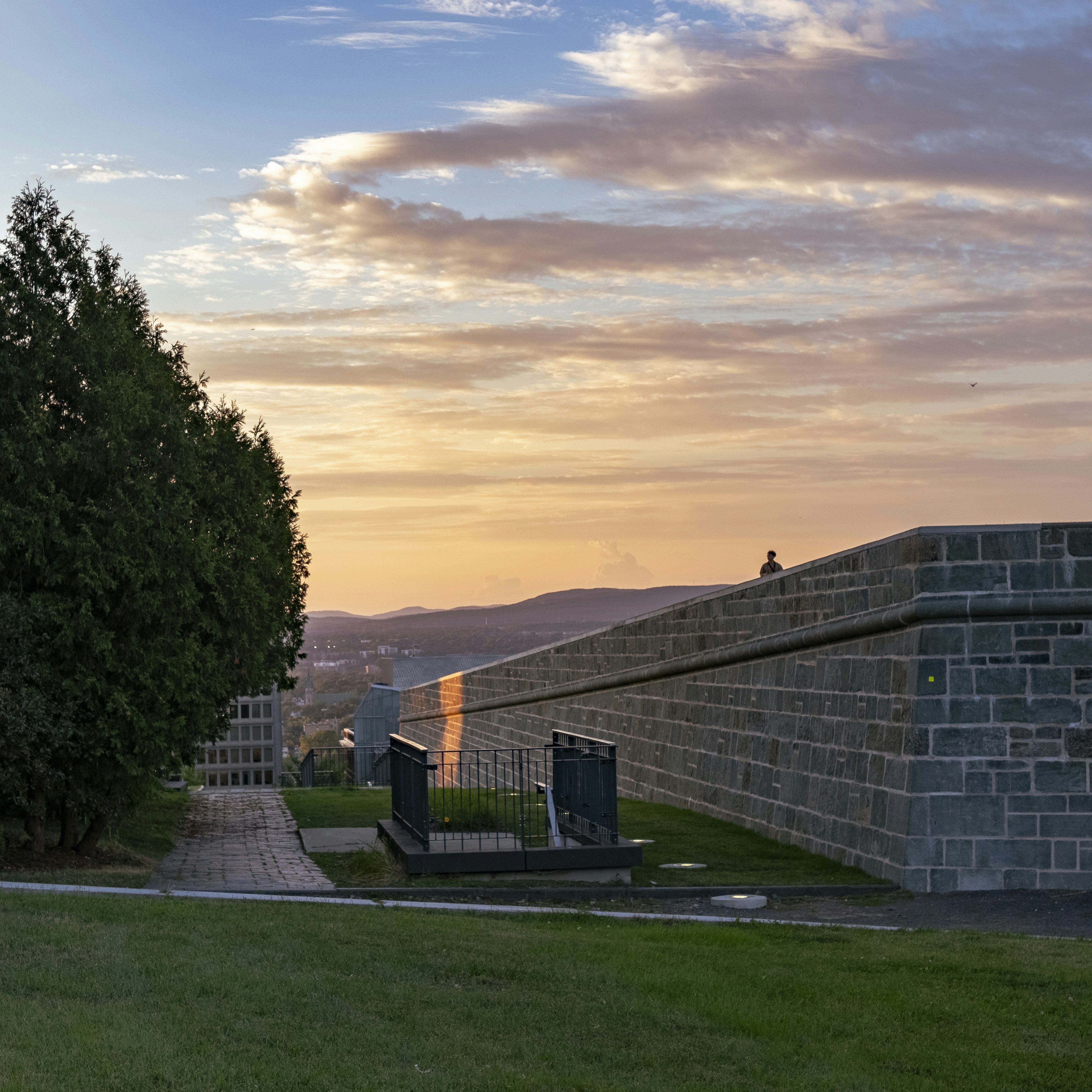 A tranquil landscape featuring a stone wall against a backdrop of a colorful sunset, with a solitary figure perched above. The scene captures the essence of peaceful reflection.