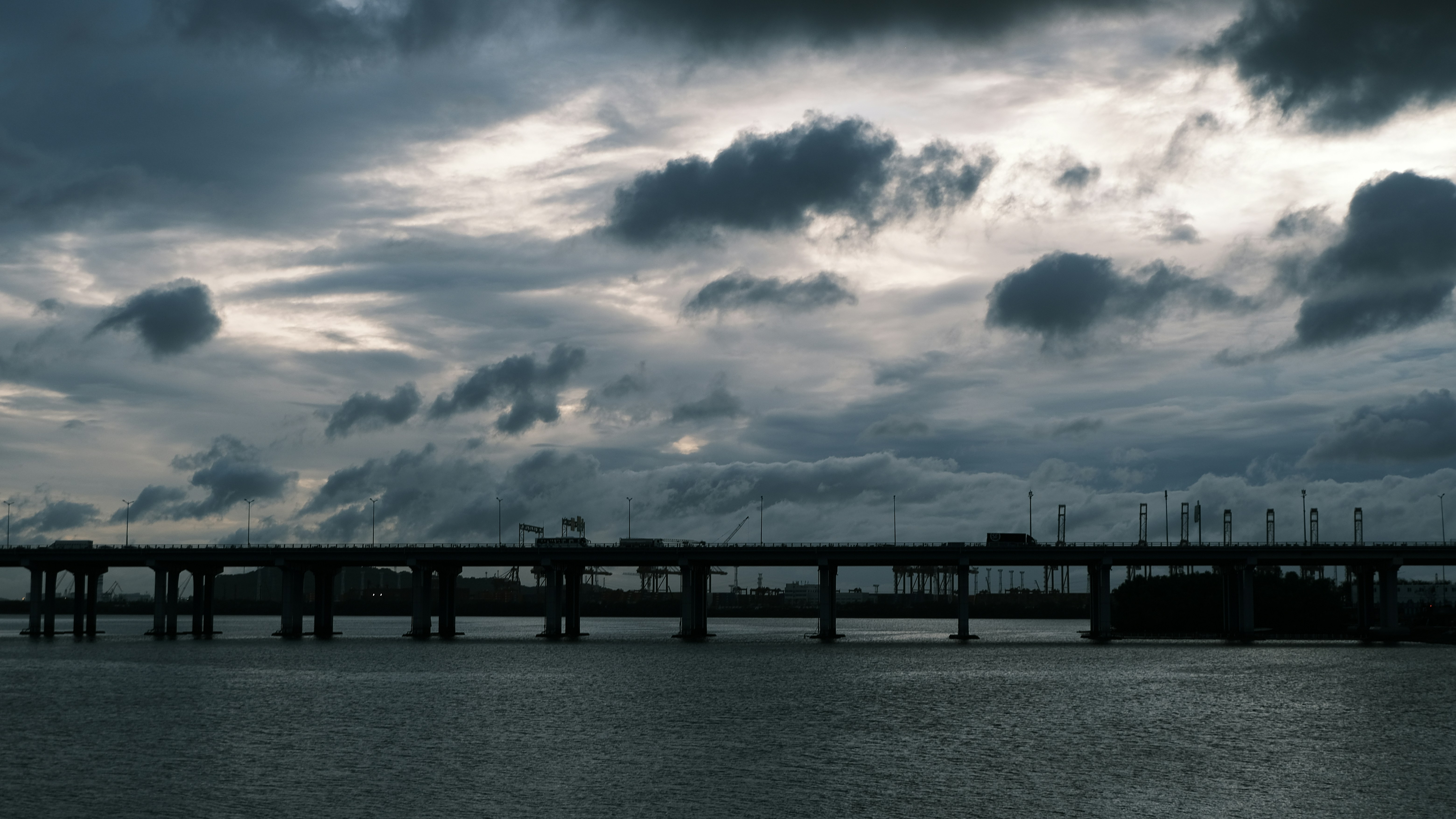 A silhouetted bridge stretches across a moody waterway under a dramatic sky filled with swirling clouds. The scene evokes a sense of tranquility amidst impending weather.