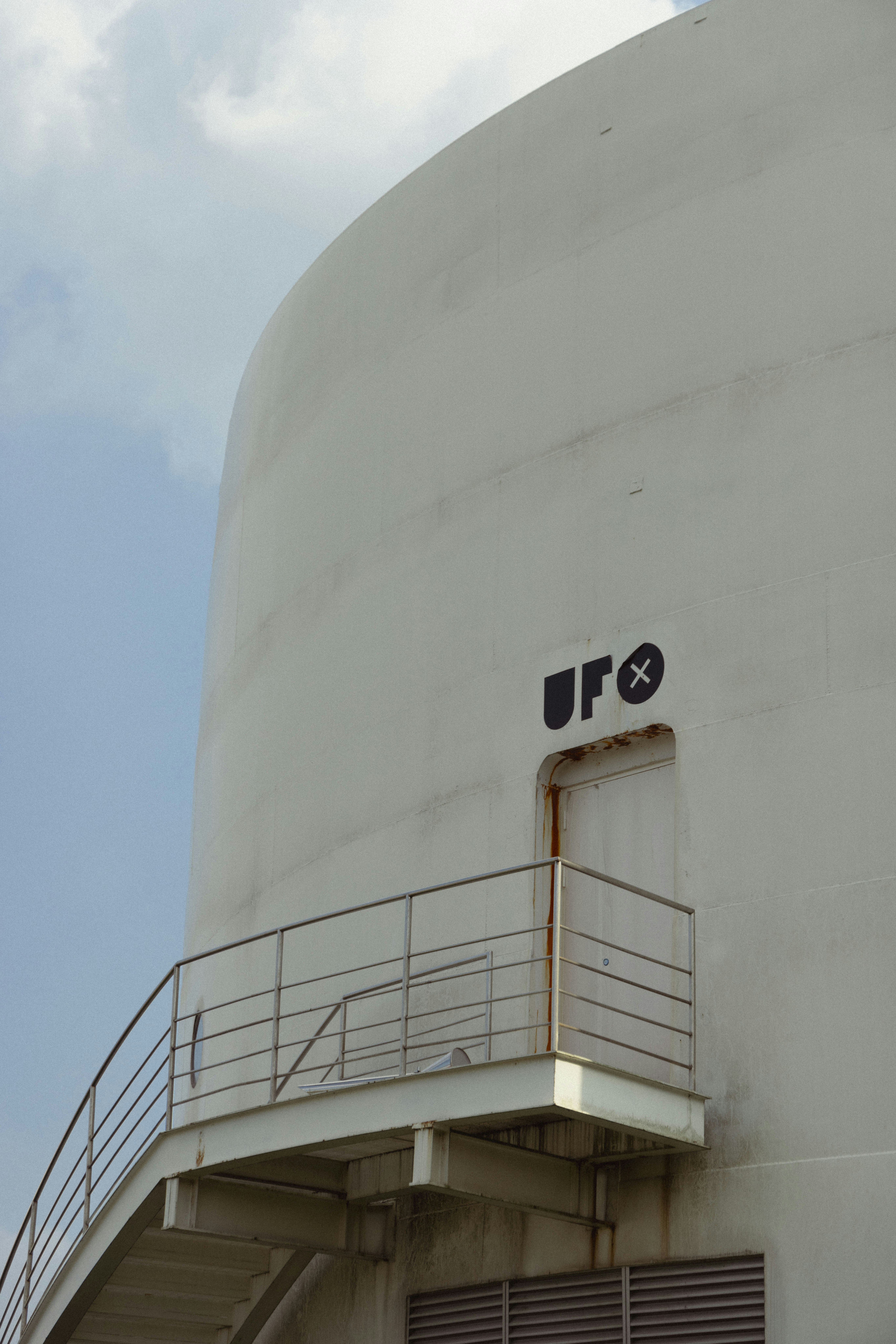 A close-up view of a cylindrical industrial structure featuring a door and a logo. The image emphasizes the interplay of lines and textures against a blue sky.