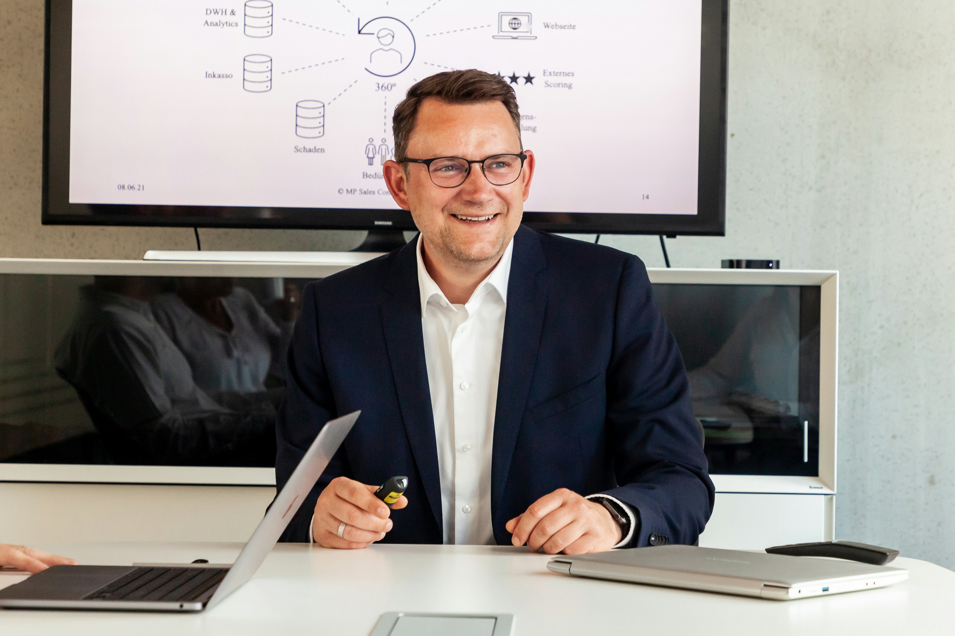 A professional man in a suit actively participates in a business meeting, surrounded by laptops and a presentation screen.