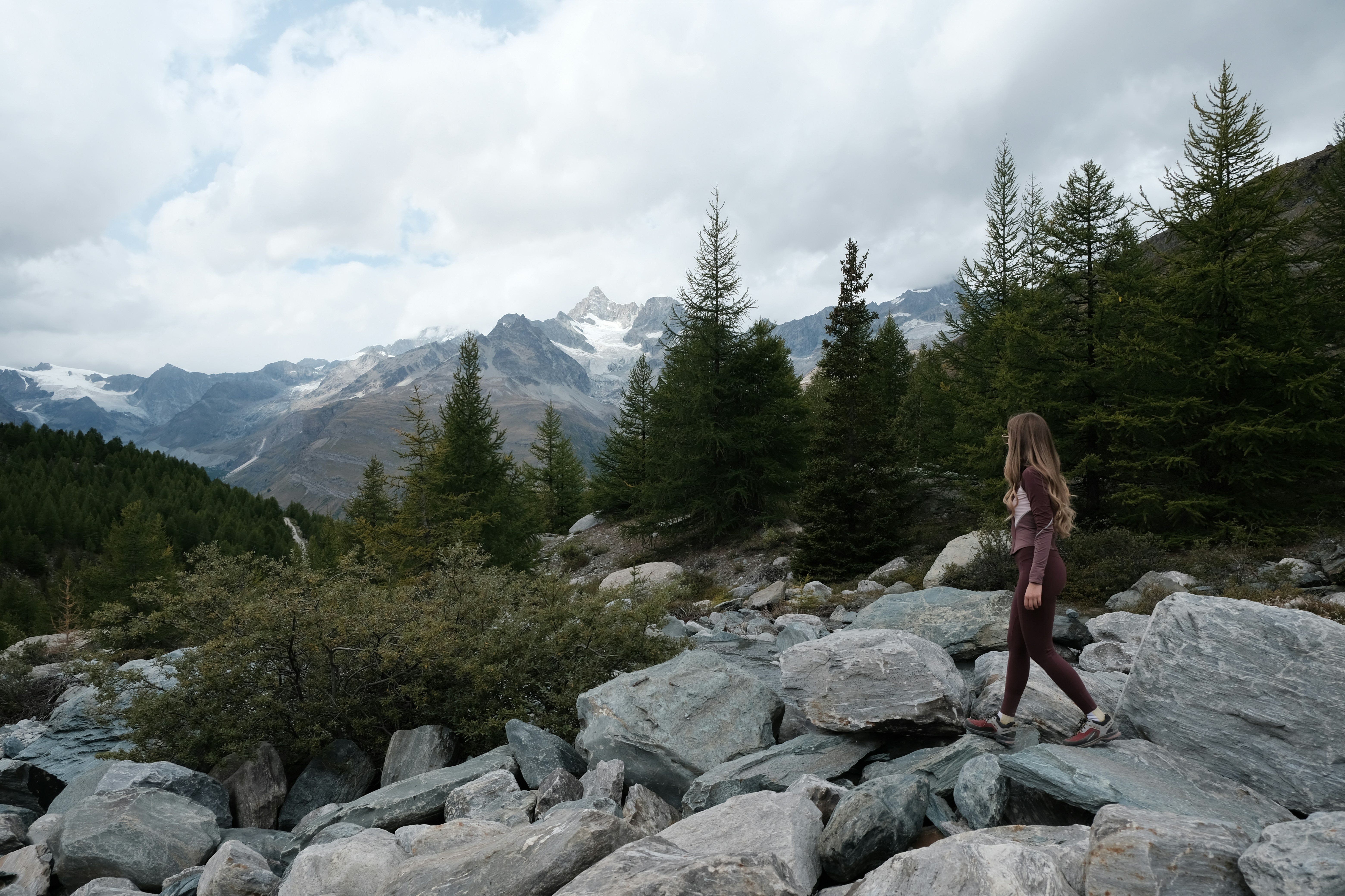 A figure traversing a rocky landscape, surrounded by lush greenery and distant snow-capped mountains under a cloudy sky.