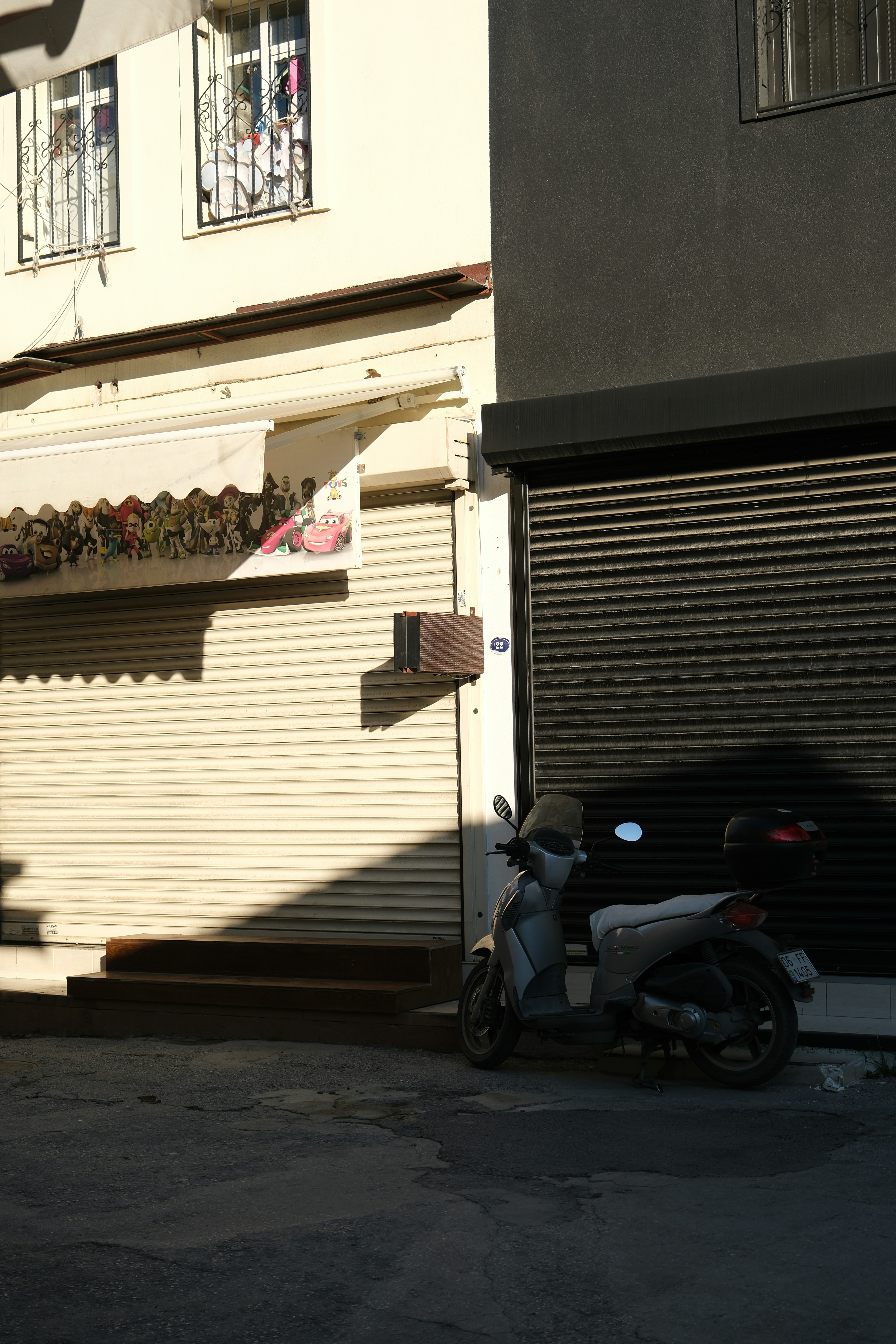 A parked scooter rests in front of closed shop shutters, one black and one white, with sharp sunlight casting strong shadows across the facade. A quiet urban street moment frozen in contrast. | A scooter parked on a street next to buildings.