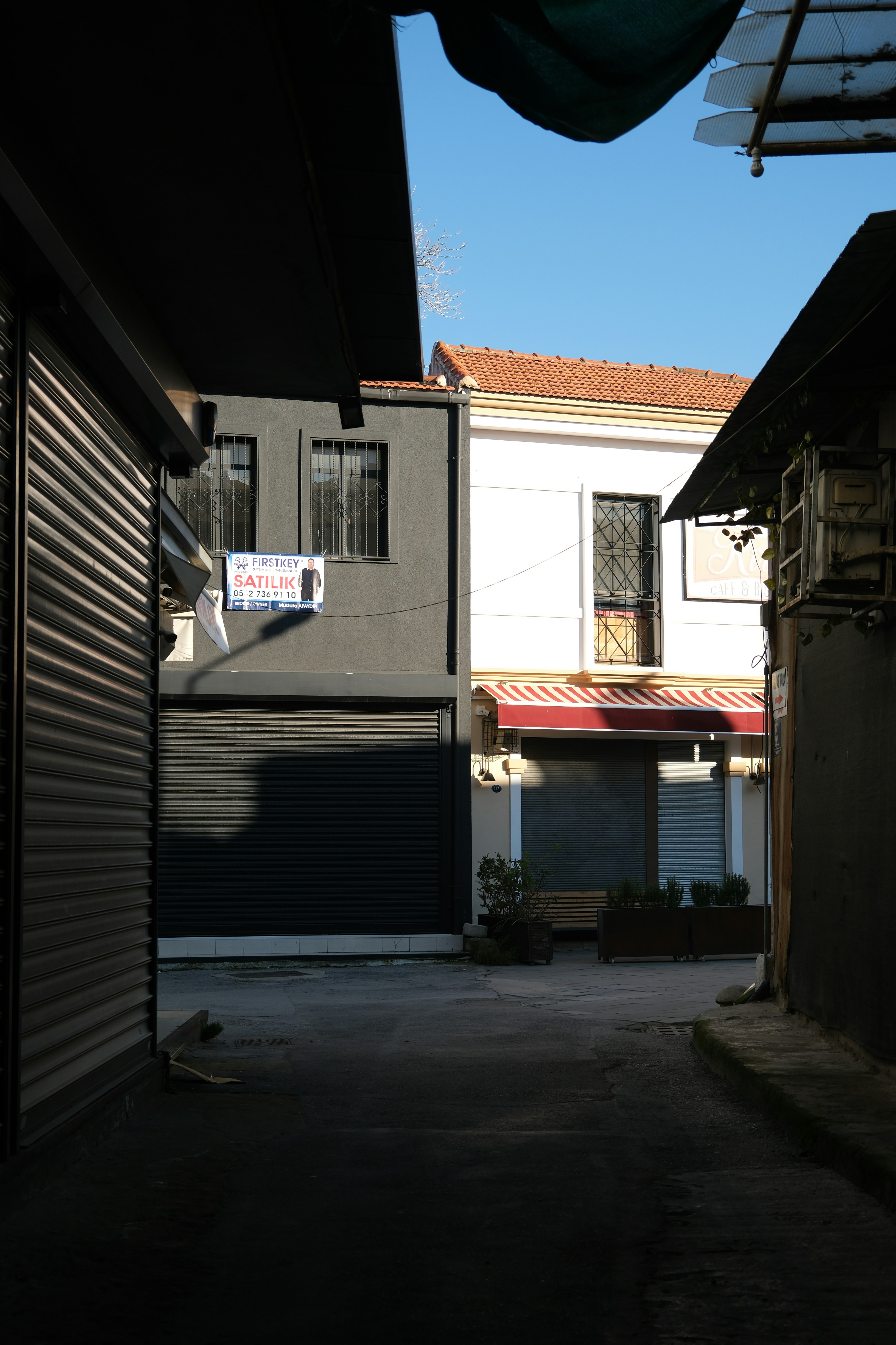 Narrow alleyway revealing contrasting buildings, with a banner indicating property for sale. The play of light and shadow enhances the urban atmosphere.