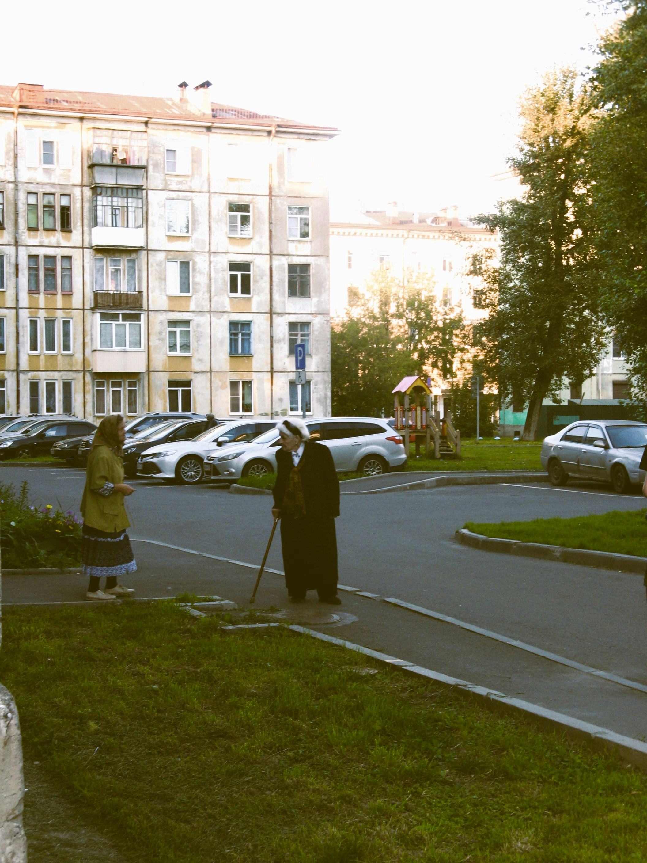 Two women engaged in conversation near a residential area, with parked cars and greenery in the background.