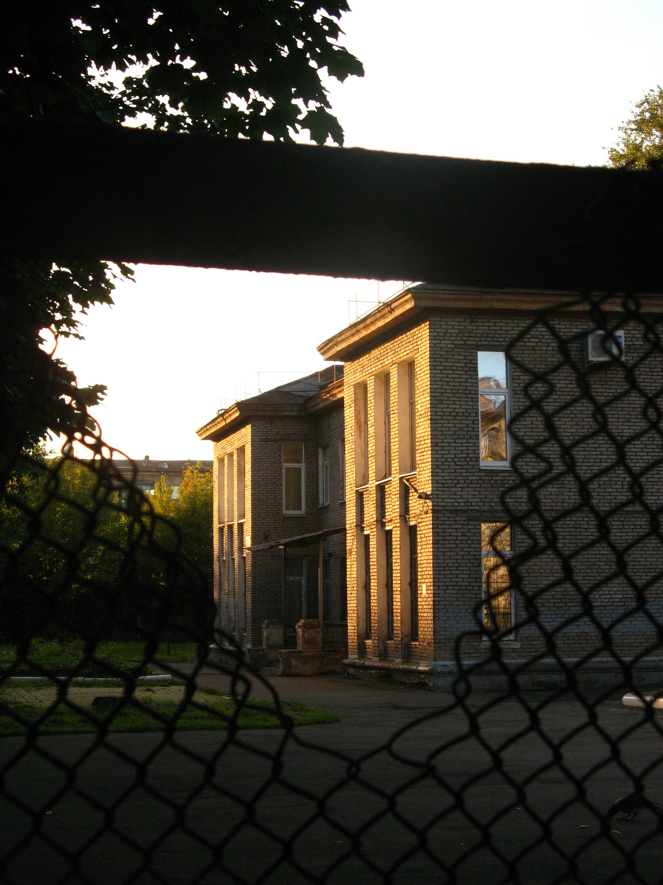 Brick building partially obscured by a fence, illuminated by warm evening light. The scene evokes a sense of nostalgia and quiet decay.