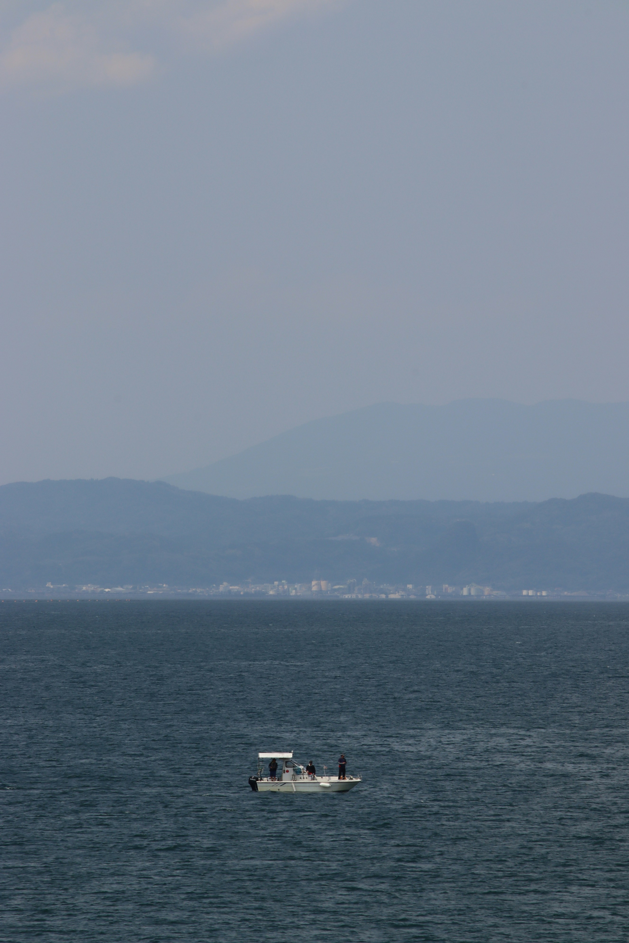 A small fishing boat with two anglers drifts peacefully on a vast ocean, framed by distant mountains and a hazy shoreline.