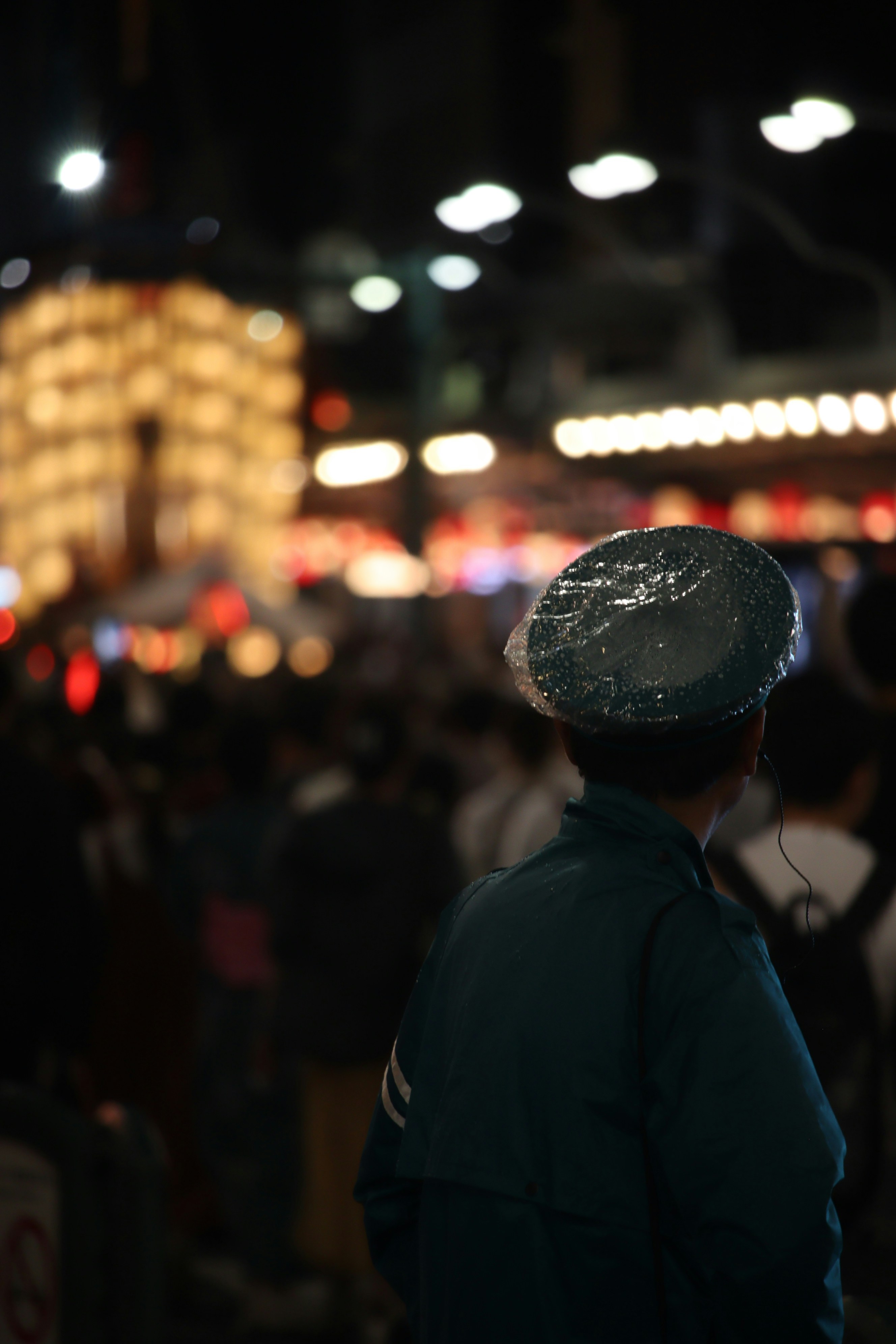 A security officer stands vigilant amid a bustling night festival, illuminated by vibrant lights and a sea of people. The atmosphere is filled with excitement and anticipation.