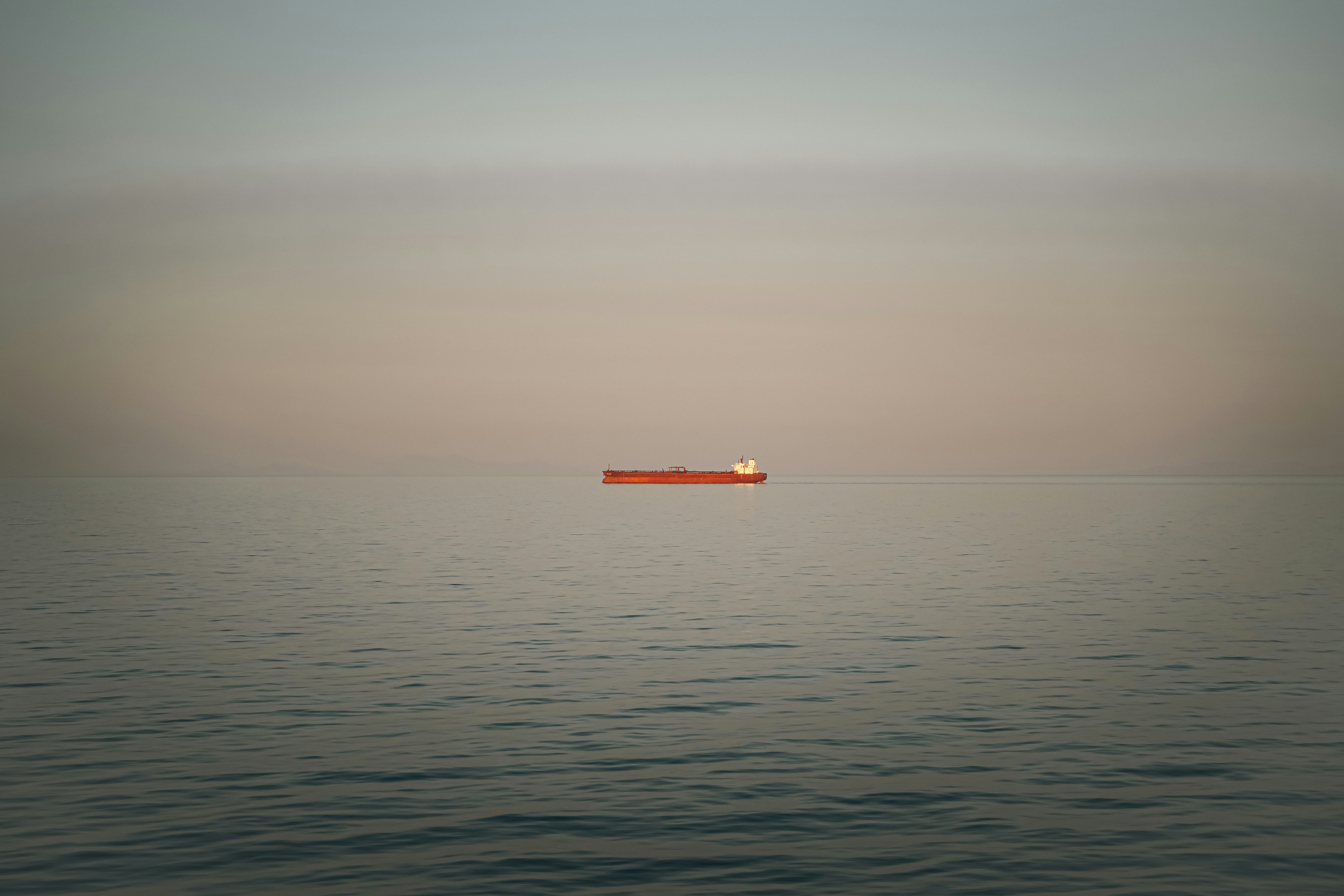 A lone cargo ship floats on calm waters under a hazy, pastel sky — captured at dusk, where the horizon nearly disappears and the sea and sky blend into quiet symmetry. | A lone cargo ship sails on a calm, vast ocean.