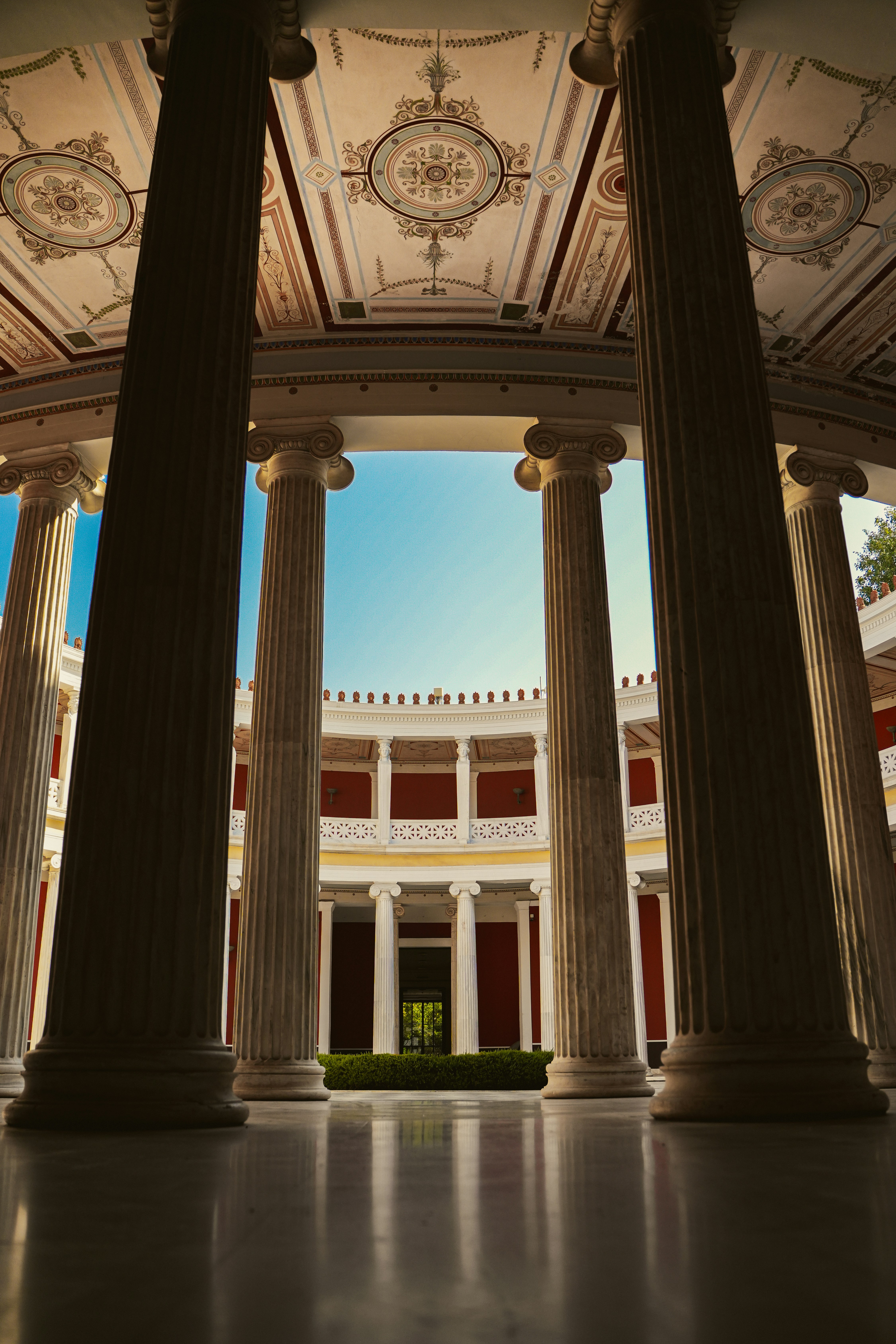 A view from within the colonnaded rotunda of Zappeion Hall in Athens, Greece — showcasing the building’s neoclassical architecture, ornate painted ceiling, and symmetrical marble columns that frame the bright Athenian sky above. | Grand columns framing a clear blue sky.