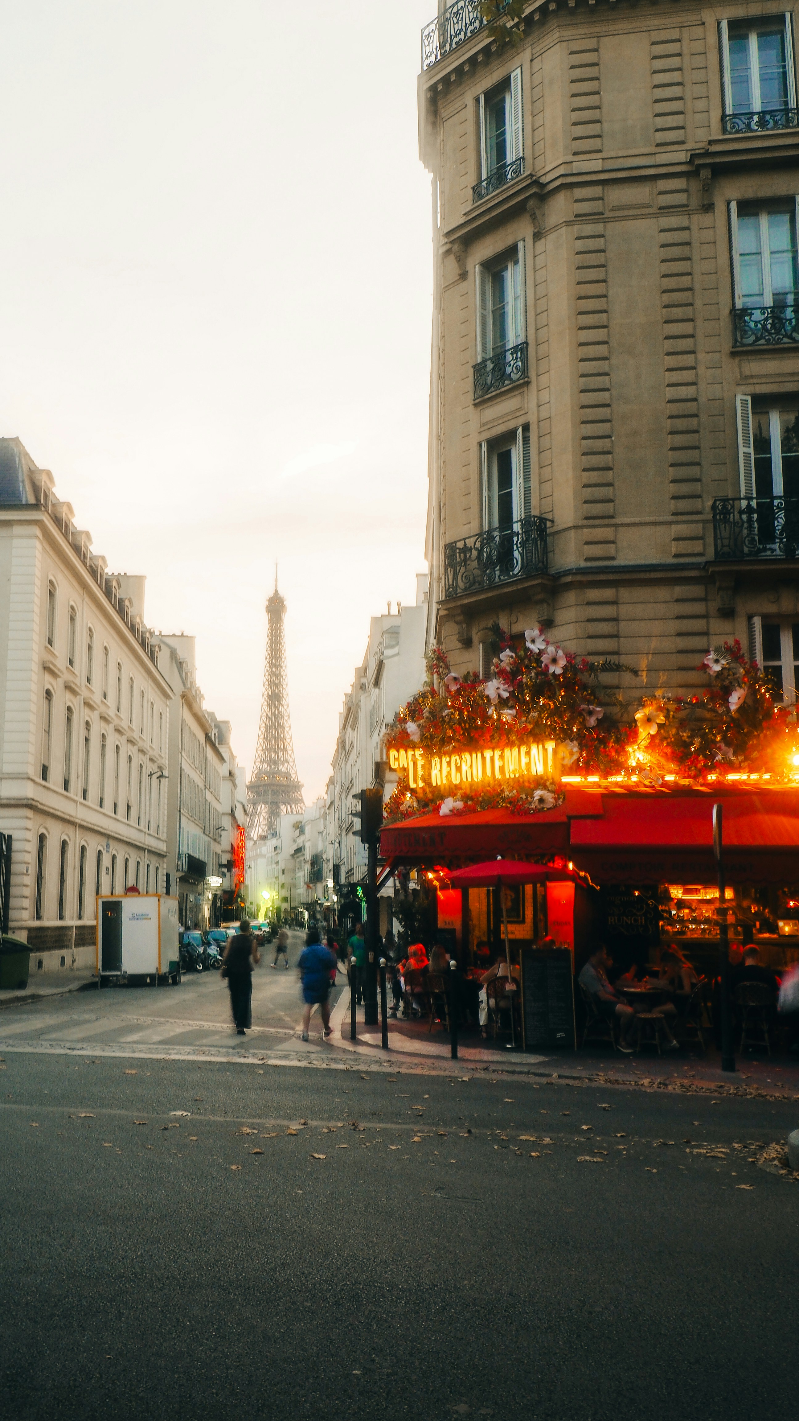 Charming Paris street scene featuring a vibrant café with illuminated signage and the Eiffel Tower in the background, evoking a warm evening ambiance.